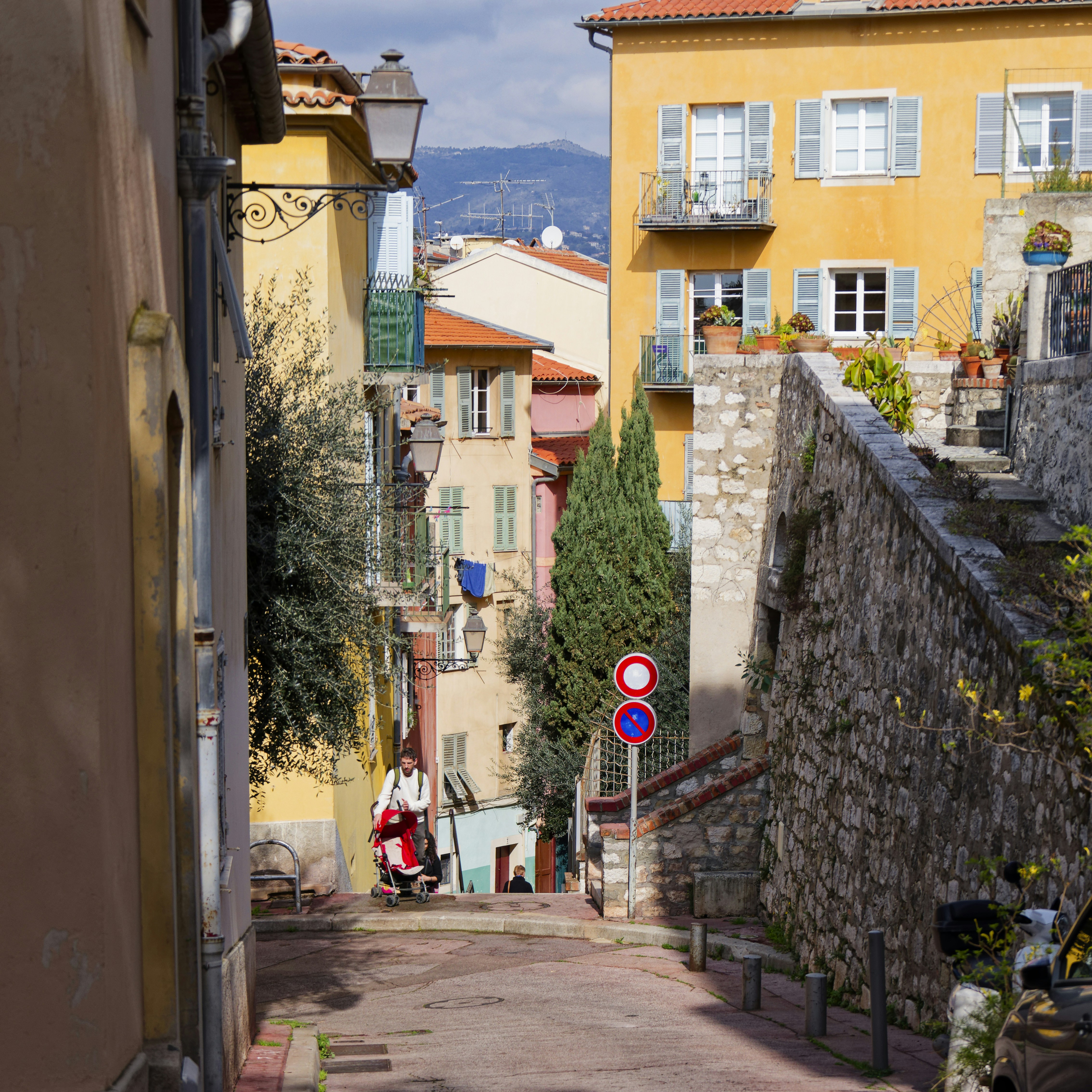 Sunlit hillside street in a colorful Mediterranean town with pastel buildings and a narrow pedestrian path. A couple with a stroller appears near the lower left, while stone walls and potted plants line the route.