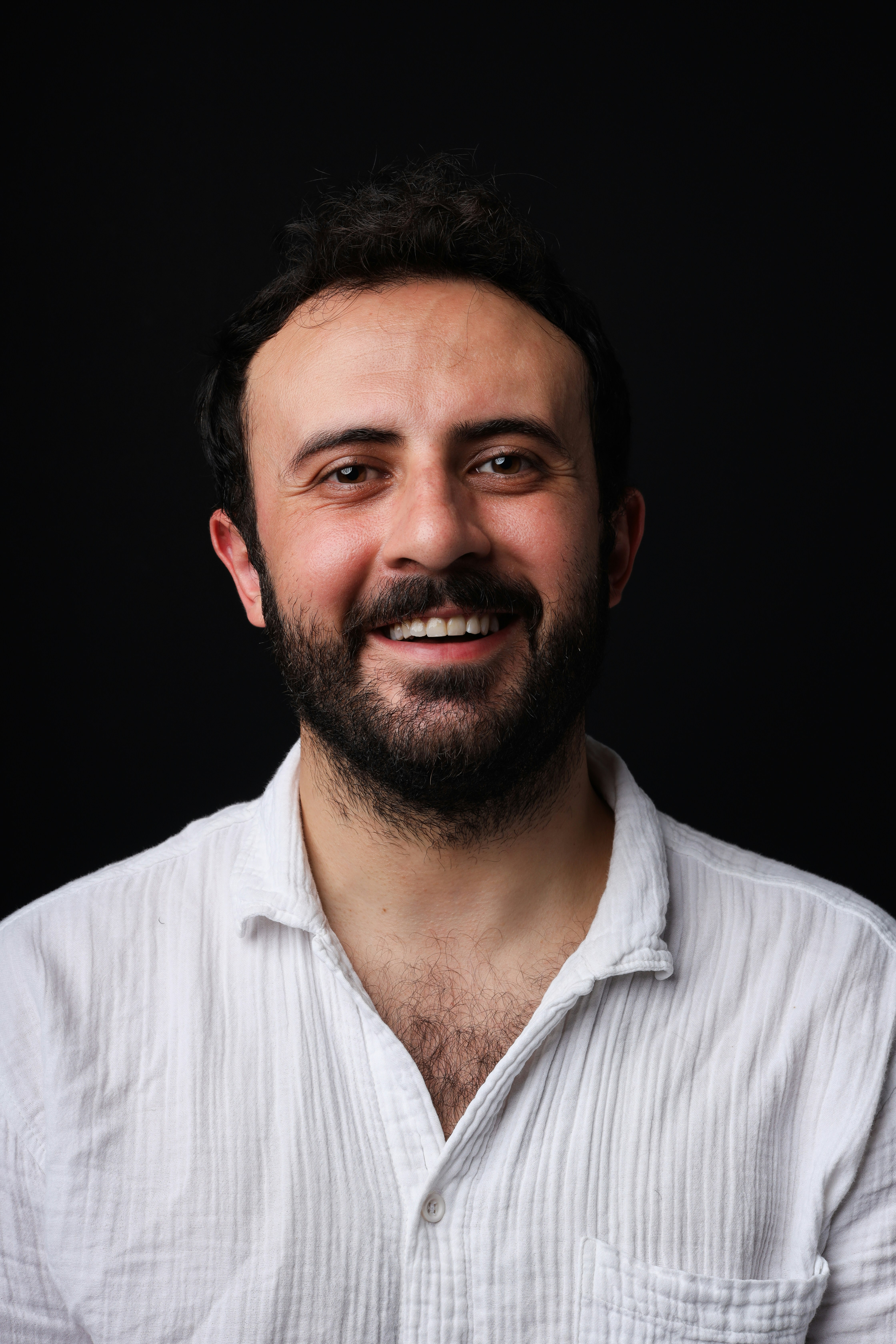 Smiling man with a beard in a white linen shirt stands against a dark backdrop, portrait-focused composition. Note: this is a photographic head-and-shoulders shot.
