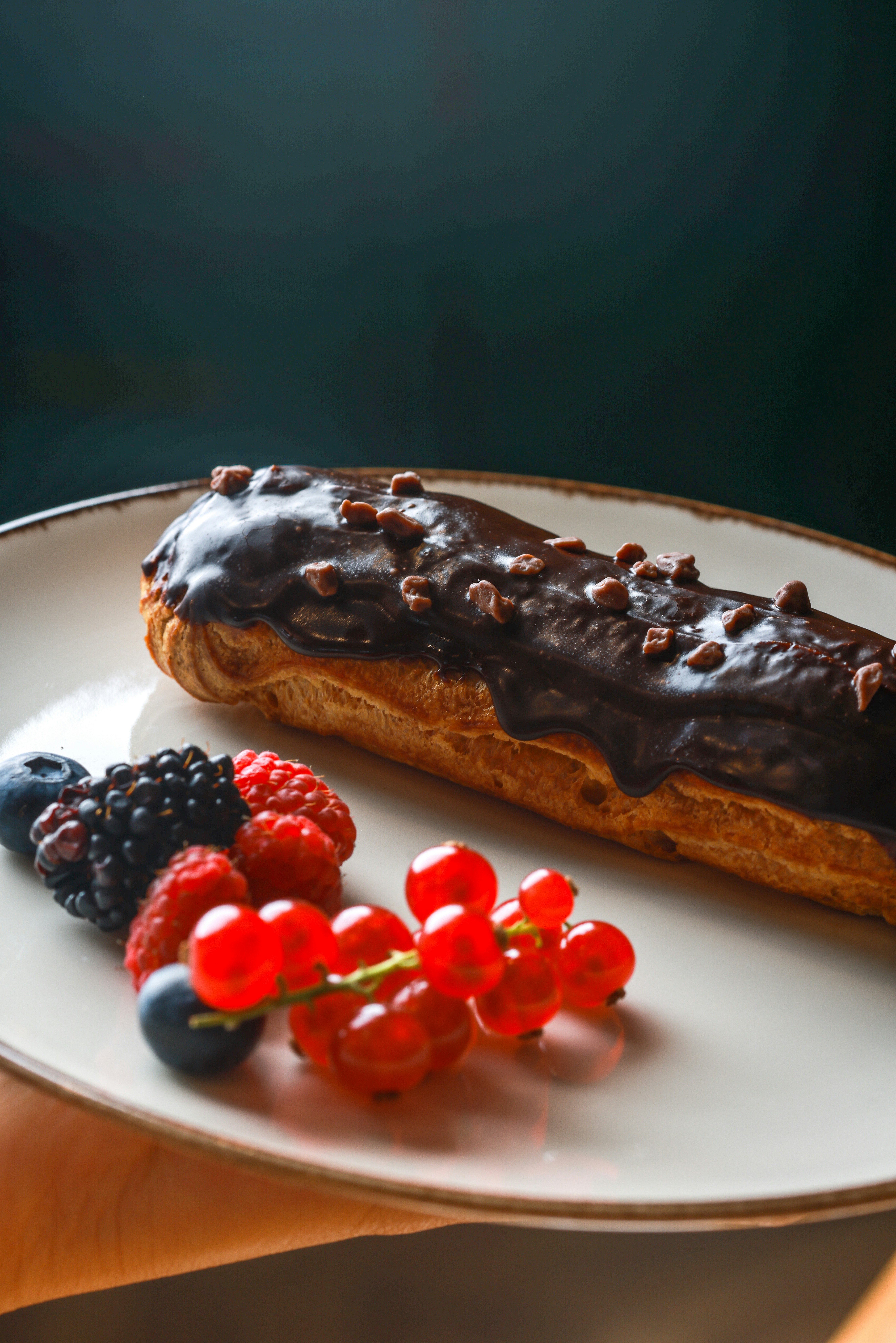 Chocolate-topped éclair plated with red currants, raspberries and blueberries on a white dish; shallow depth of field emphasizes the pastry while dark teal background provides contrast.