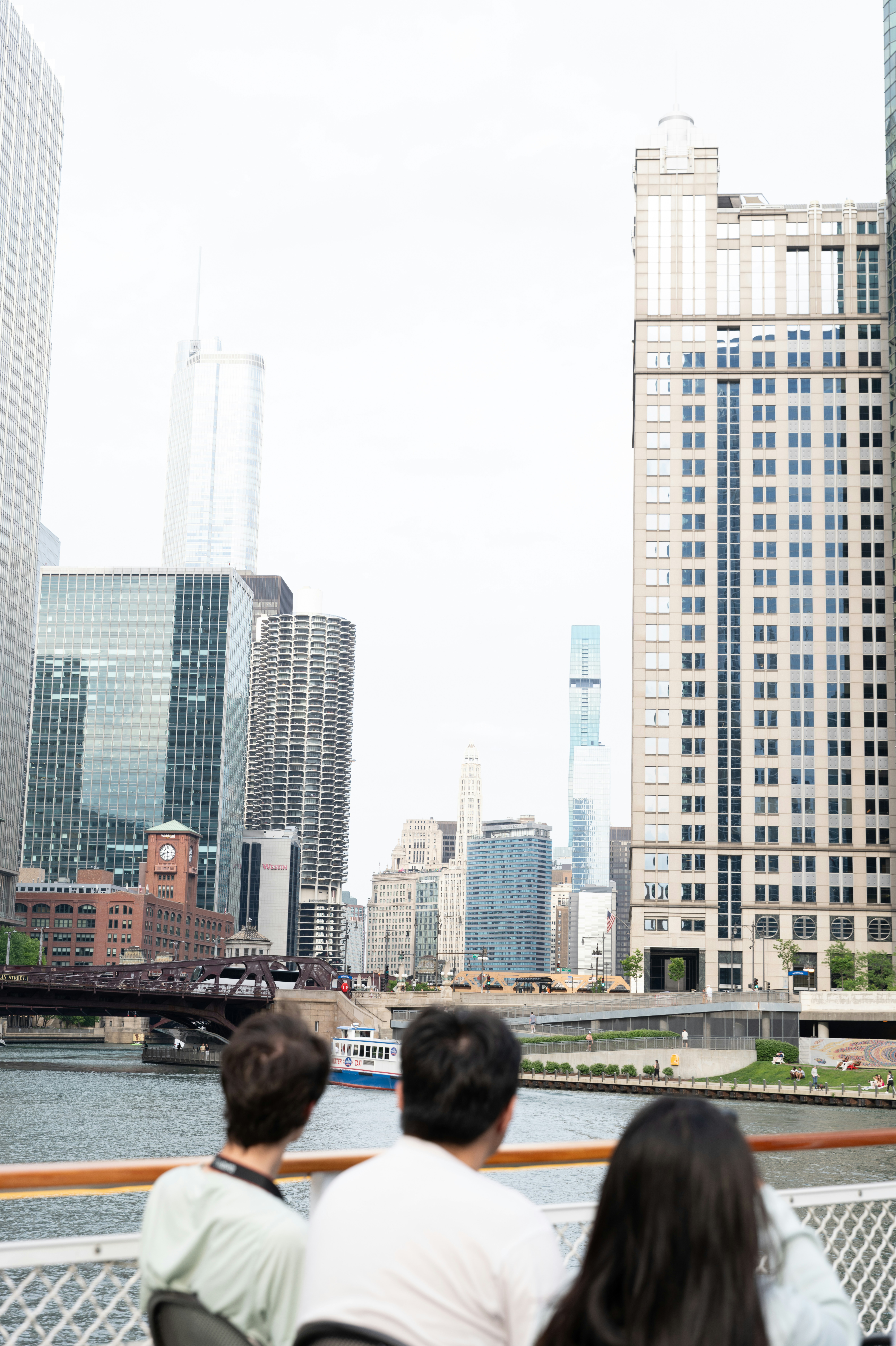 Three people in the foreground overlook a calm river with a modern cityscape of glass towers in the background.