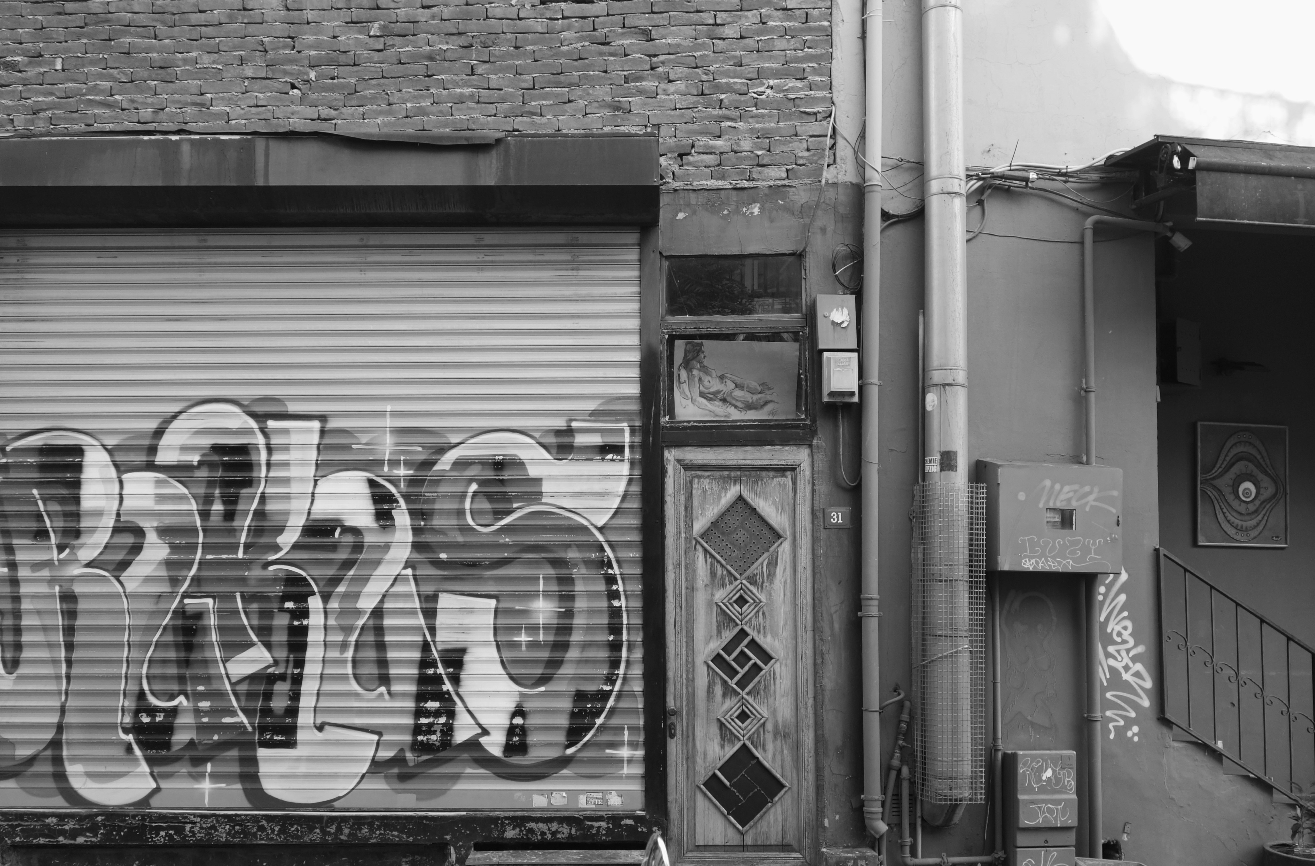 Graffiti-covered rolling shutter and adjacent wall with weathered doors and utility fixtures; a quiet street corner captured in monochrome to emphasize texture and urban decay.