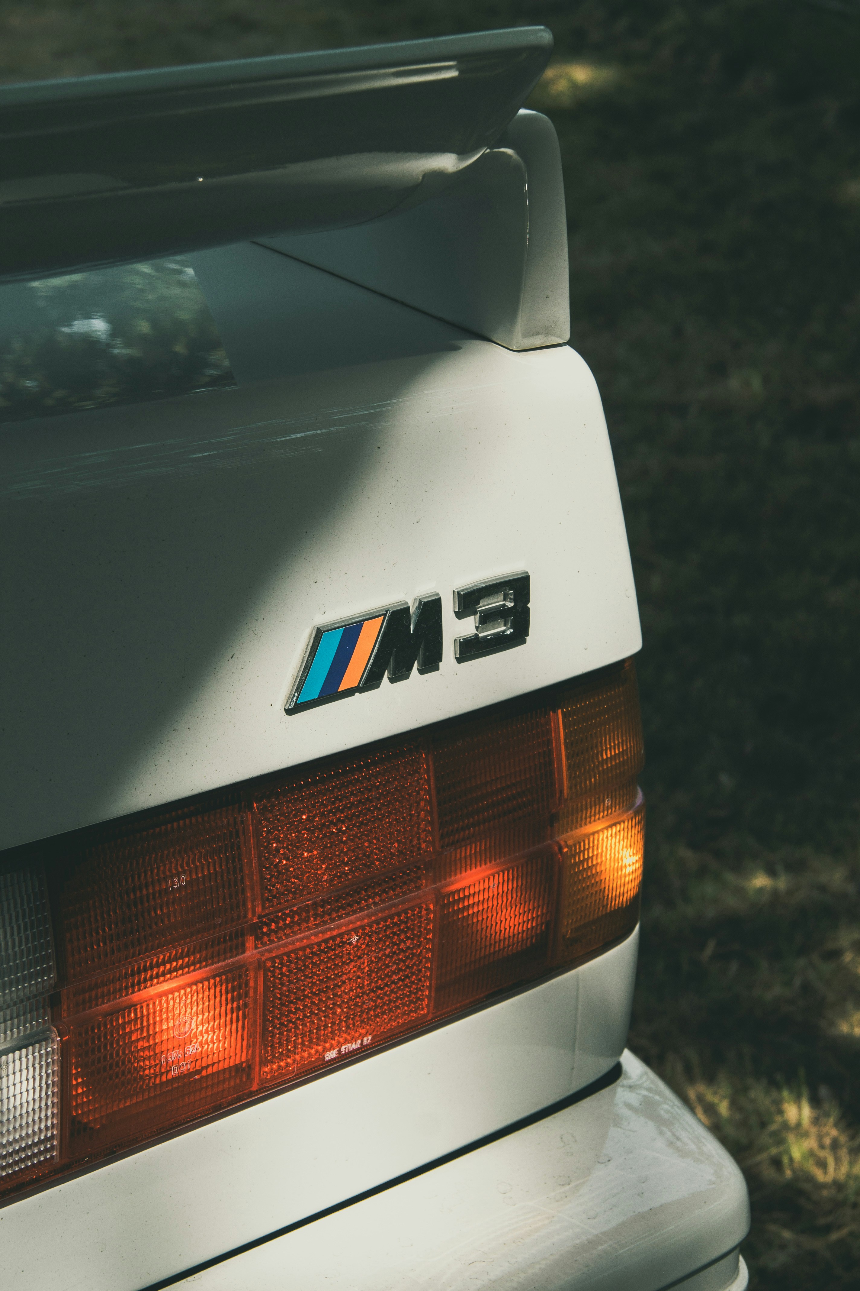 Close-up of the rear of a white BMW M3, highlighting the M3 badge and tail light assembly with a natural outdoor backdrop.