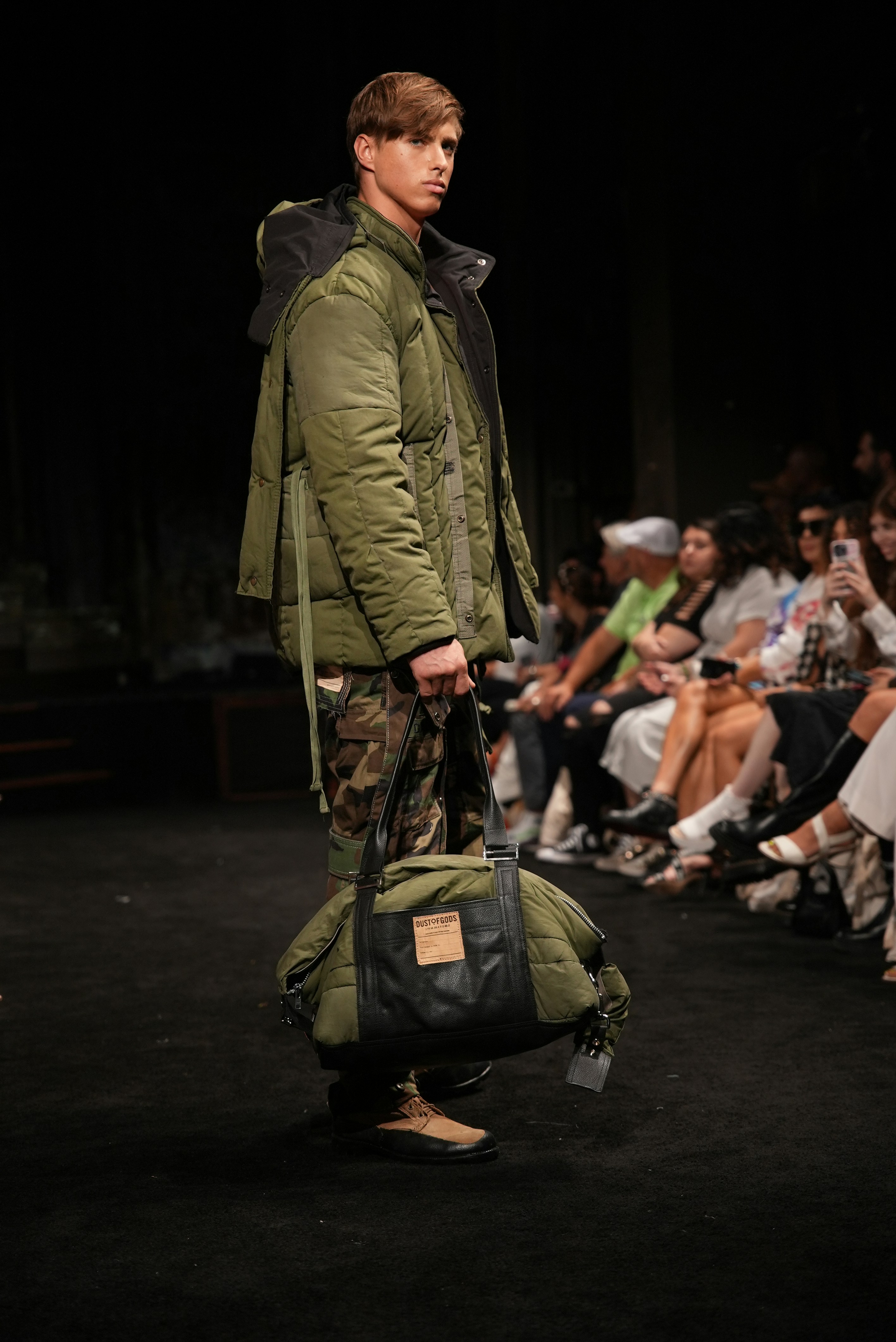 Model in a khaki green padded jacket walks the runway carrying a matching duffel-style bag; audience sits along the side under dim lights. This is a fashion show photograph.