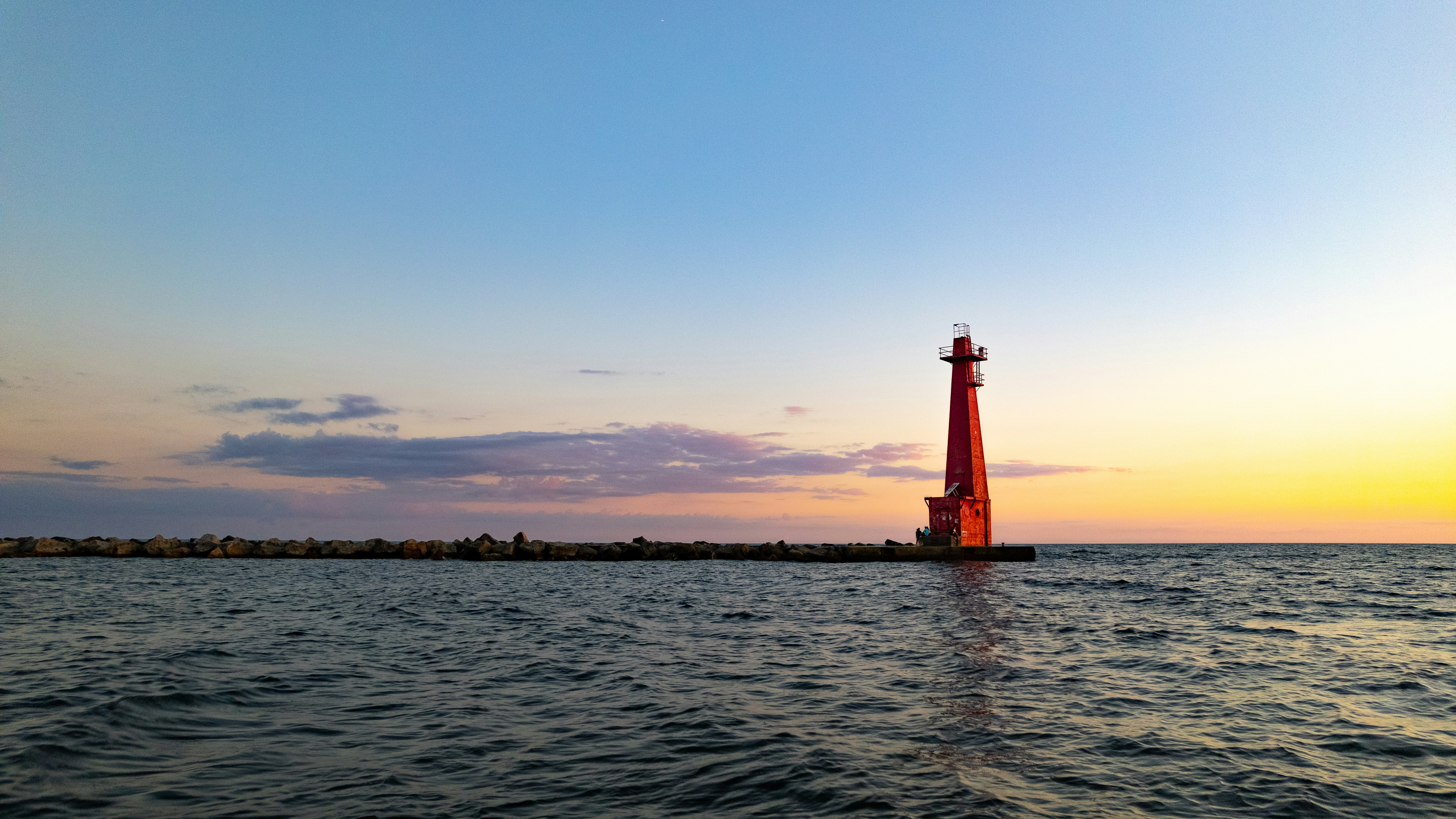Lone red lighthouse on a protective breakwater at sunset, with calm sea and soft pastel sky.
