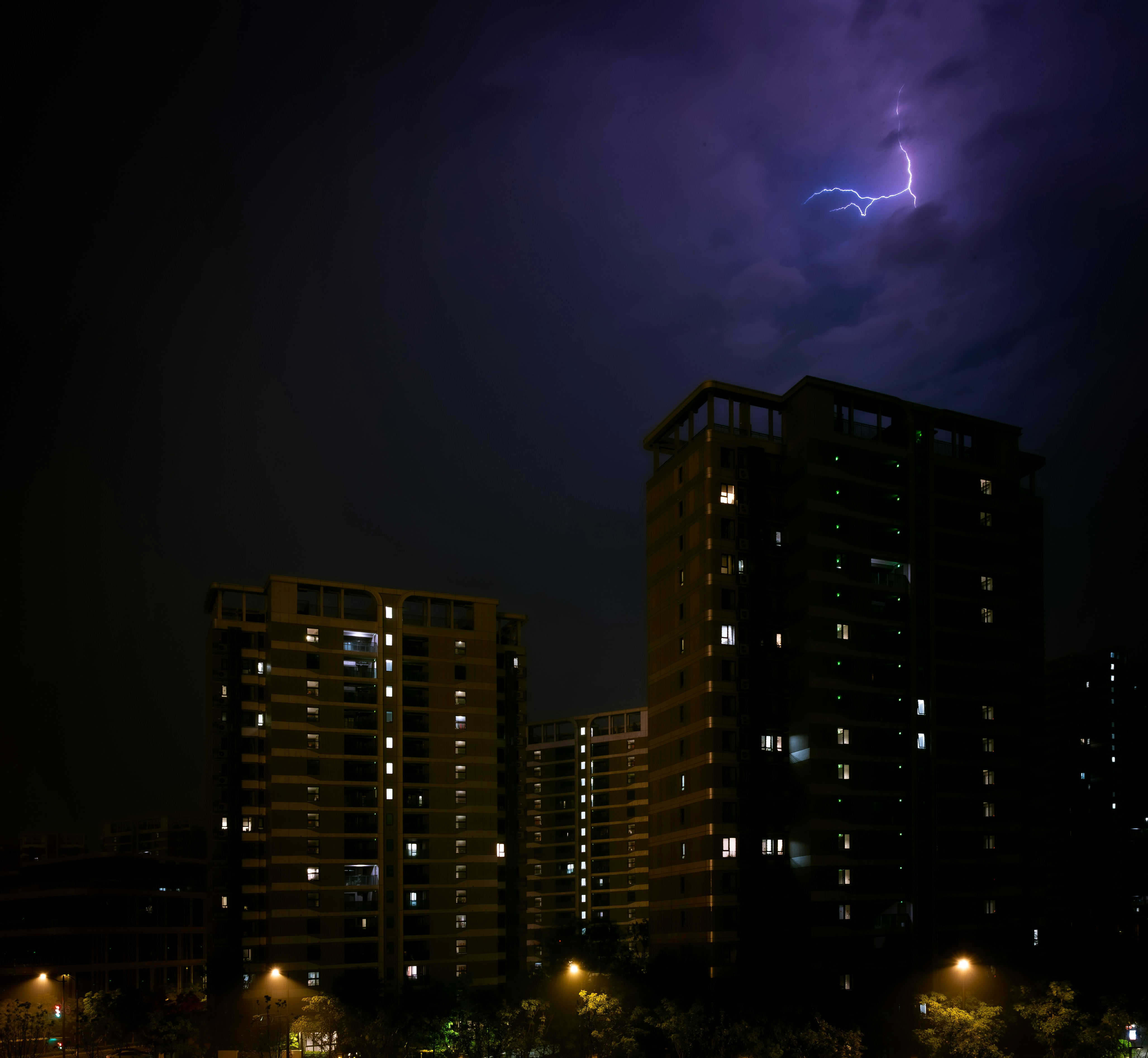 Night urban scene with lit apartment towers under a stormy sky; a bolt of lightning arcs above the buildings, adding dramatic energy to the composition. This is a photographic capture of a dark, moody cityscape.