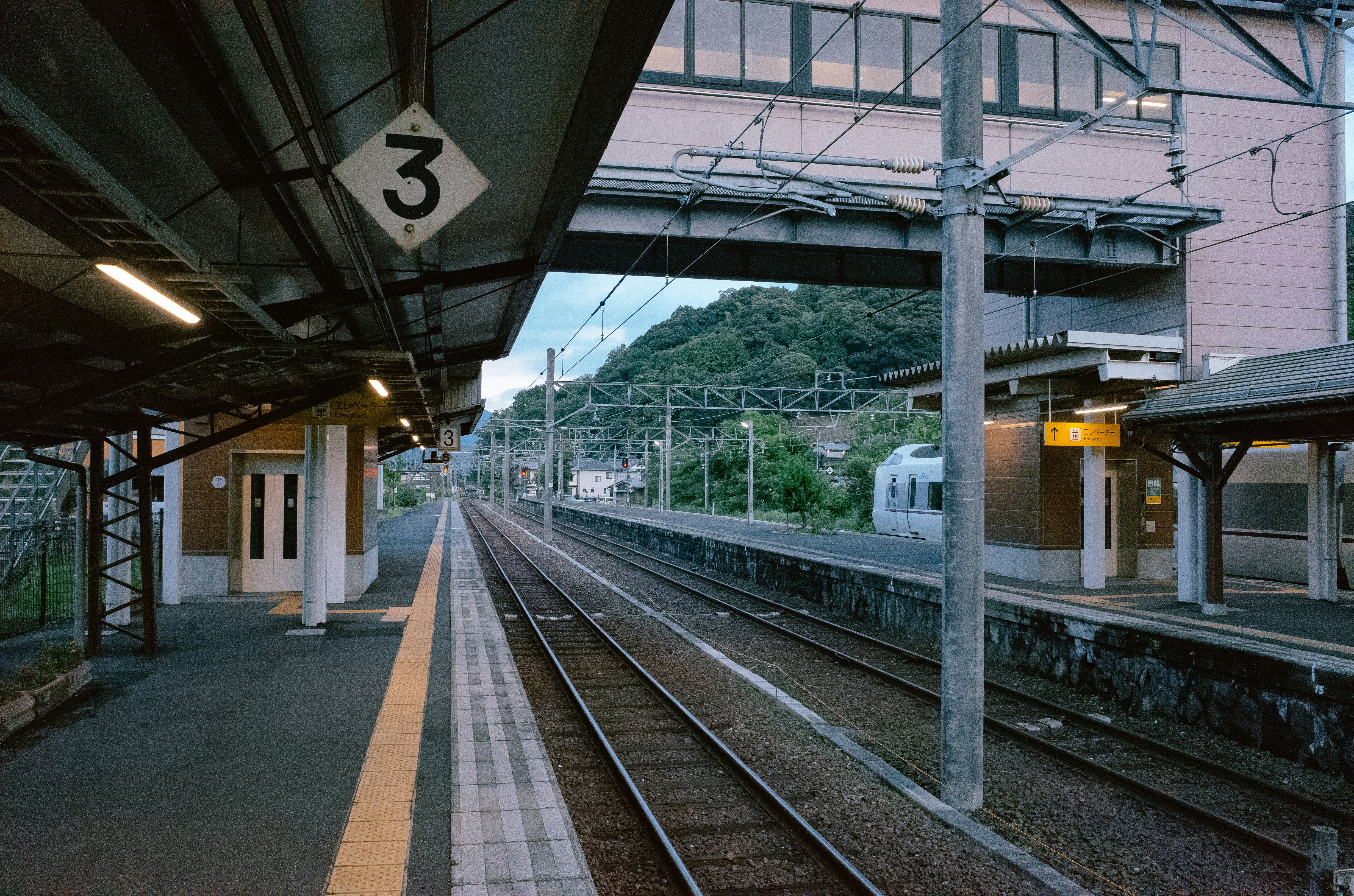 Train station platform with tracks receding toward a mountainous backdrop, captured in soft light; the image emphasizes symmetry and perspective with a distant train and clear signage.