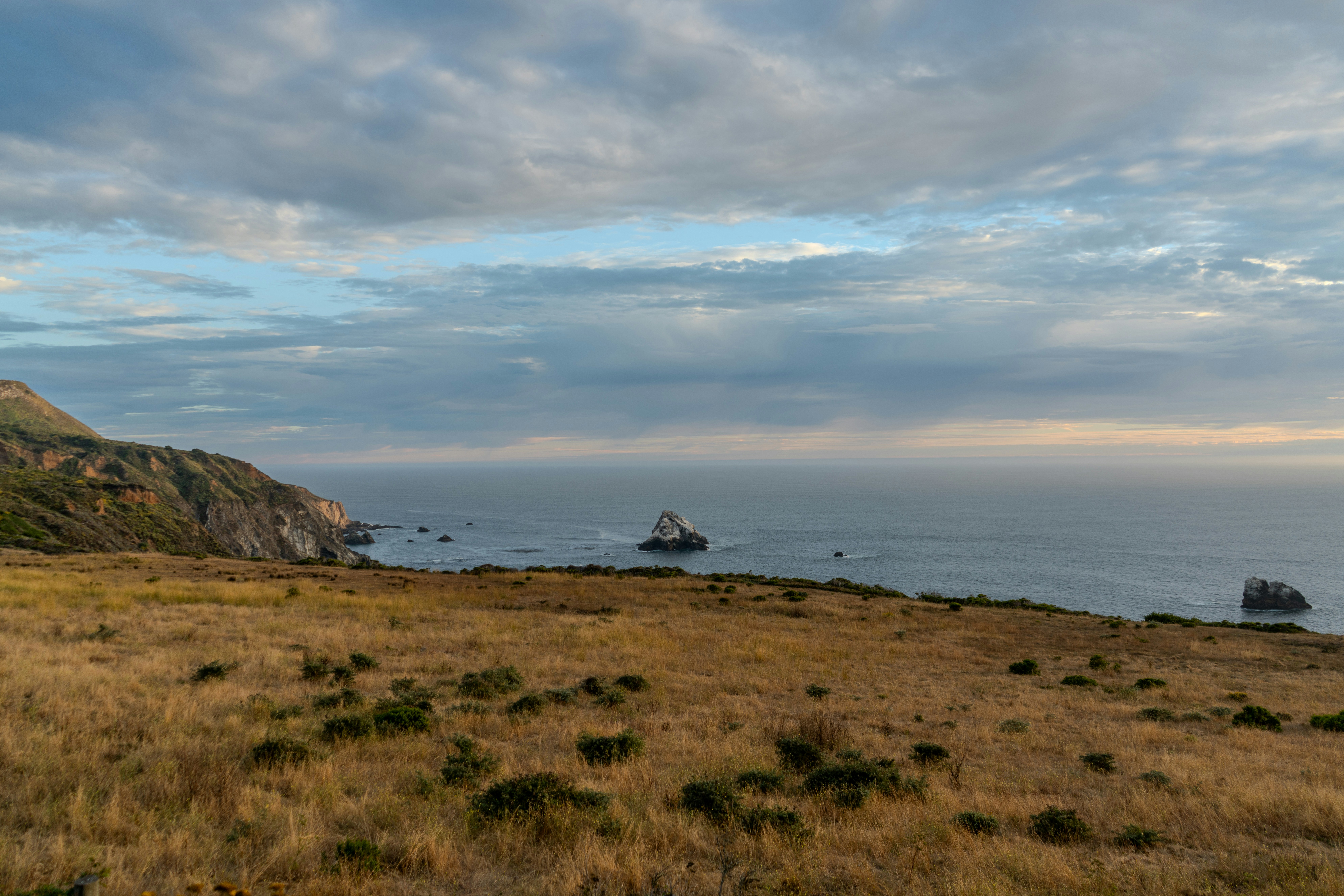 Sunlit coastal plain with dry grass extends toward rocky shoreline and calm sea under a cloudy sky.