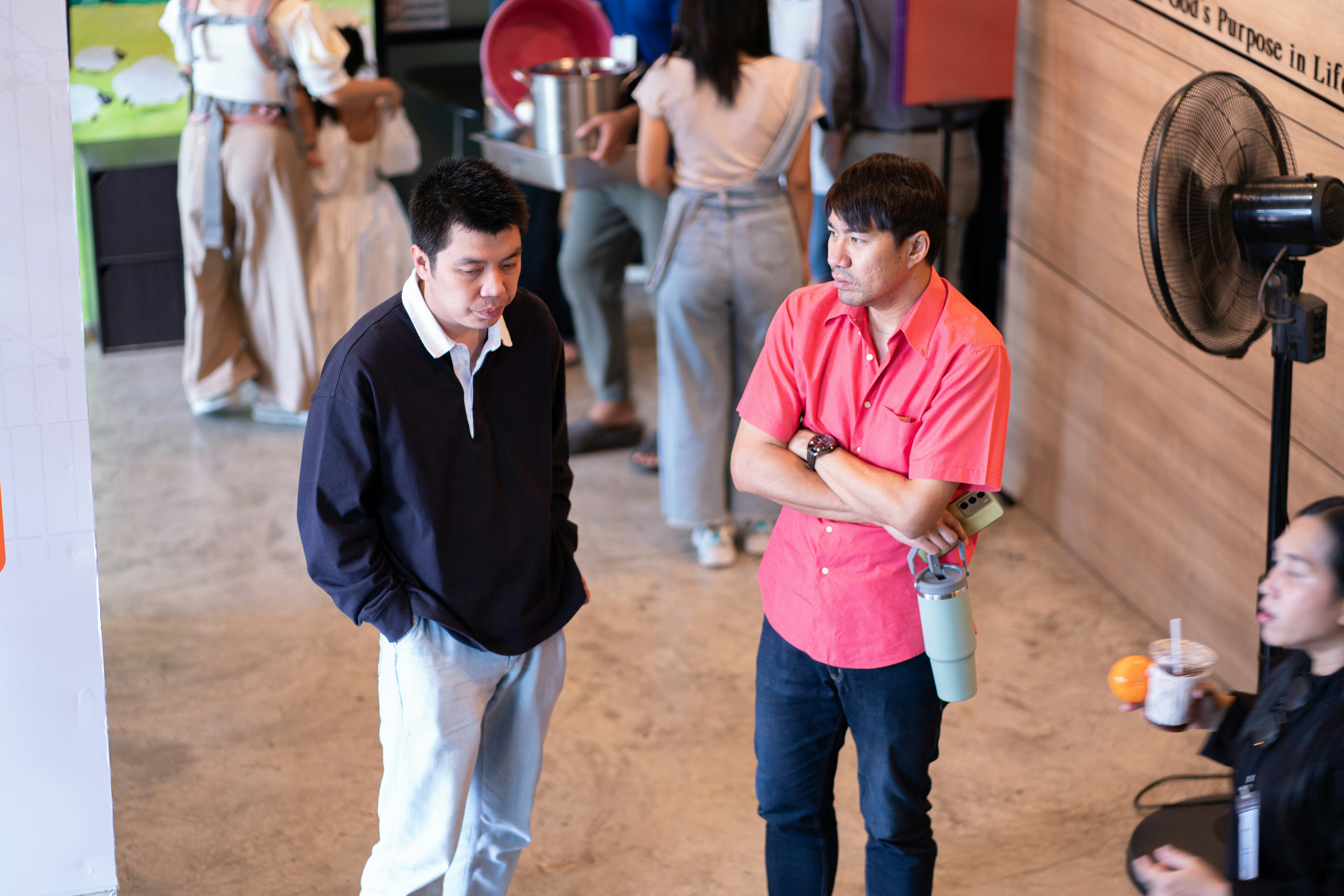 Two men in a casual showroom pause near a wooden wall and standing fan