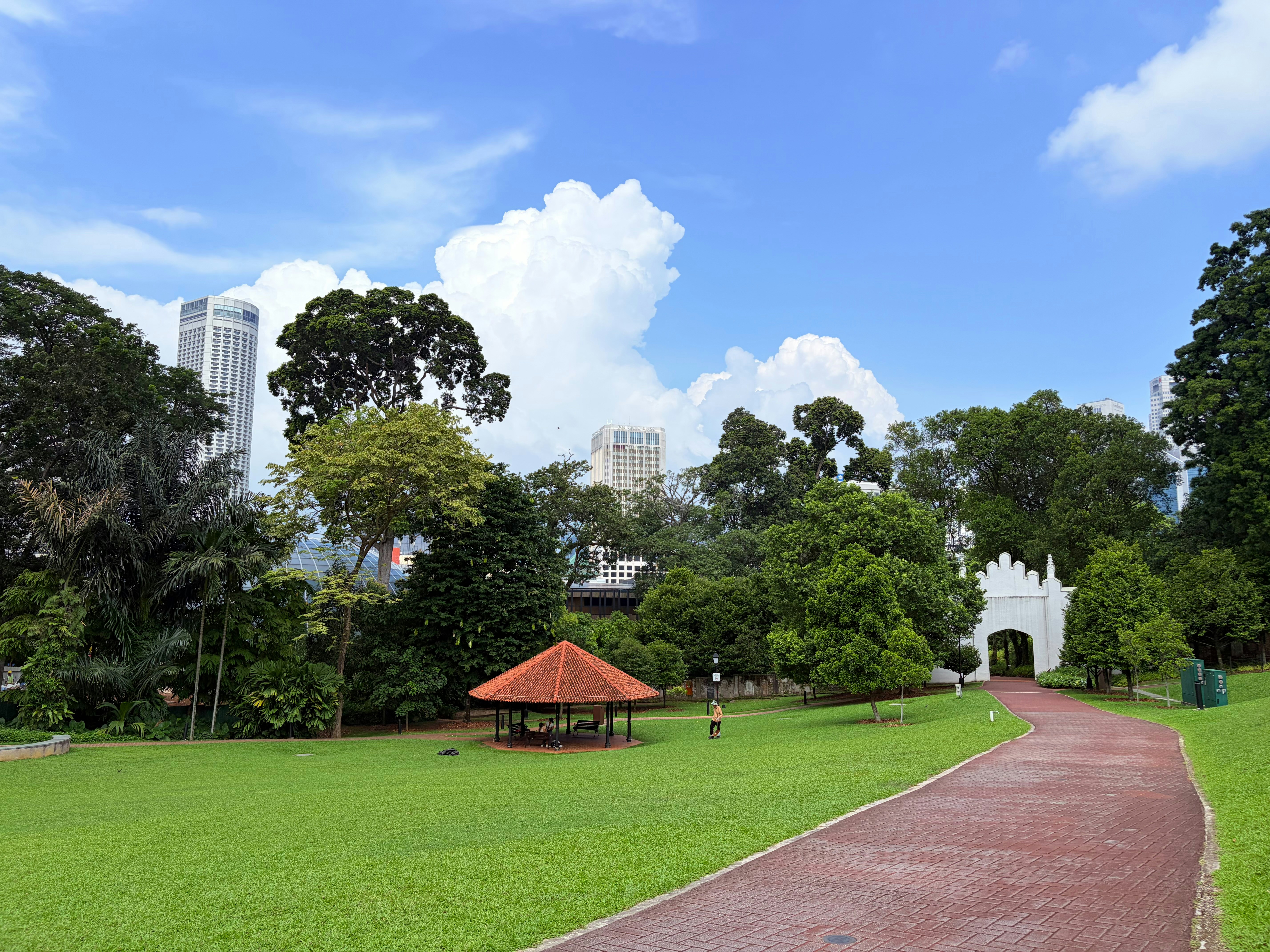 Sunlit park scene with a brick path leading to a white archway, surrounded by green trees and a small thatched gazebo on a bright day. This image is a garden/park photograph.