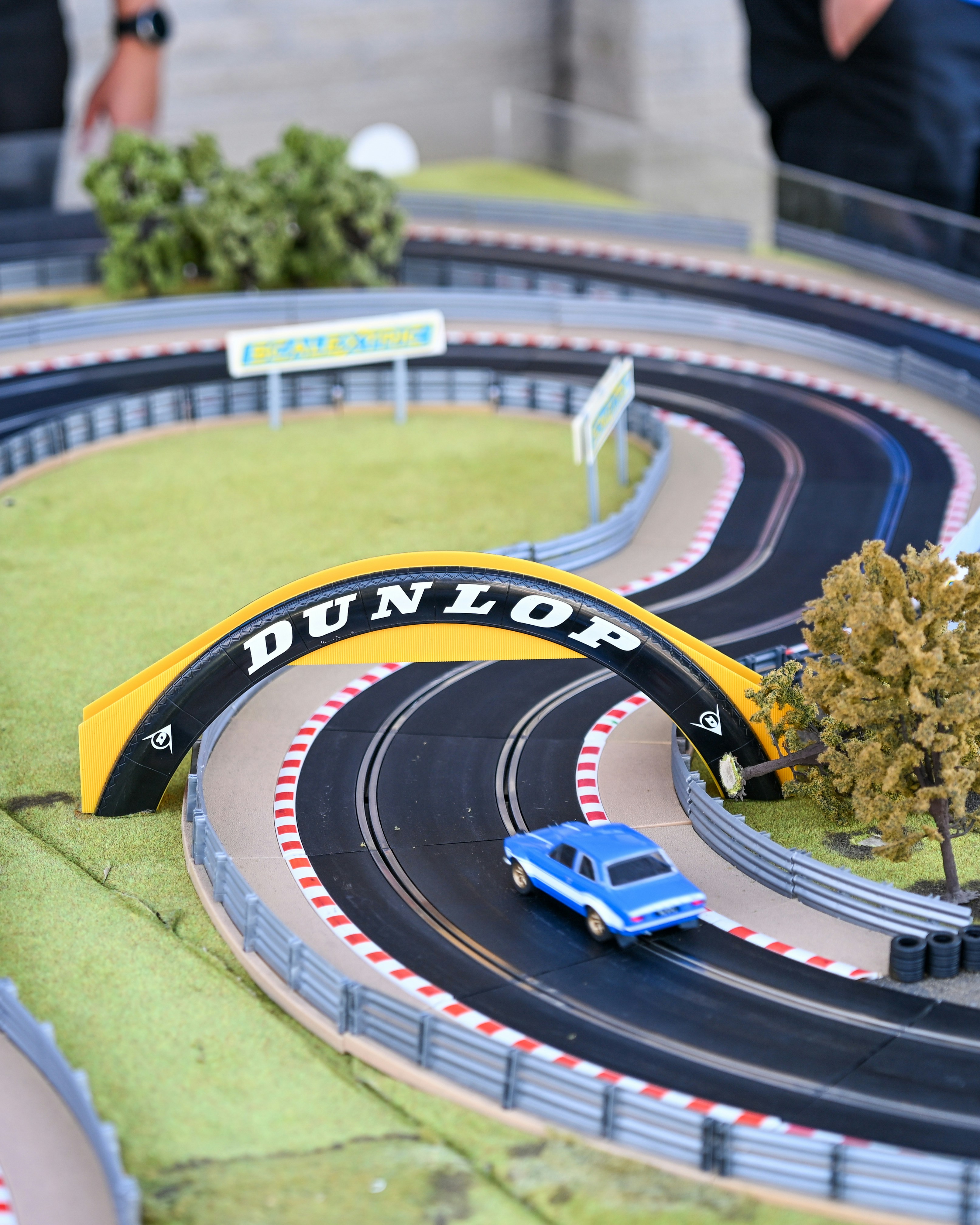 A miniature racing track features a blue car under a Dunlop arch, with winding lanes and surrounding faux grass; the scene resembles a detailed model setup.
