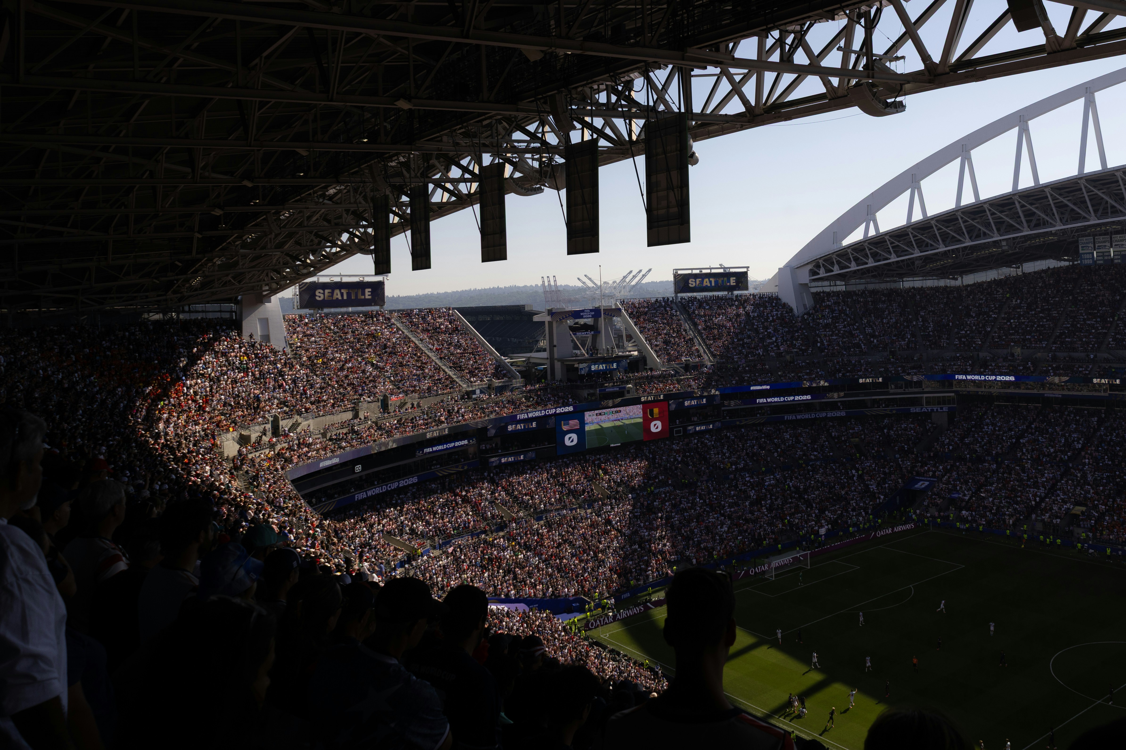 A sunlit stadium stands packed with spectators, viewed from a shaded terrace; the field and audience form a sweeping arc under a steel canopy, capturing the scale of a live match day.