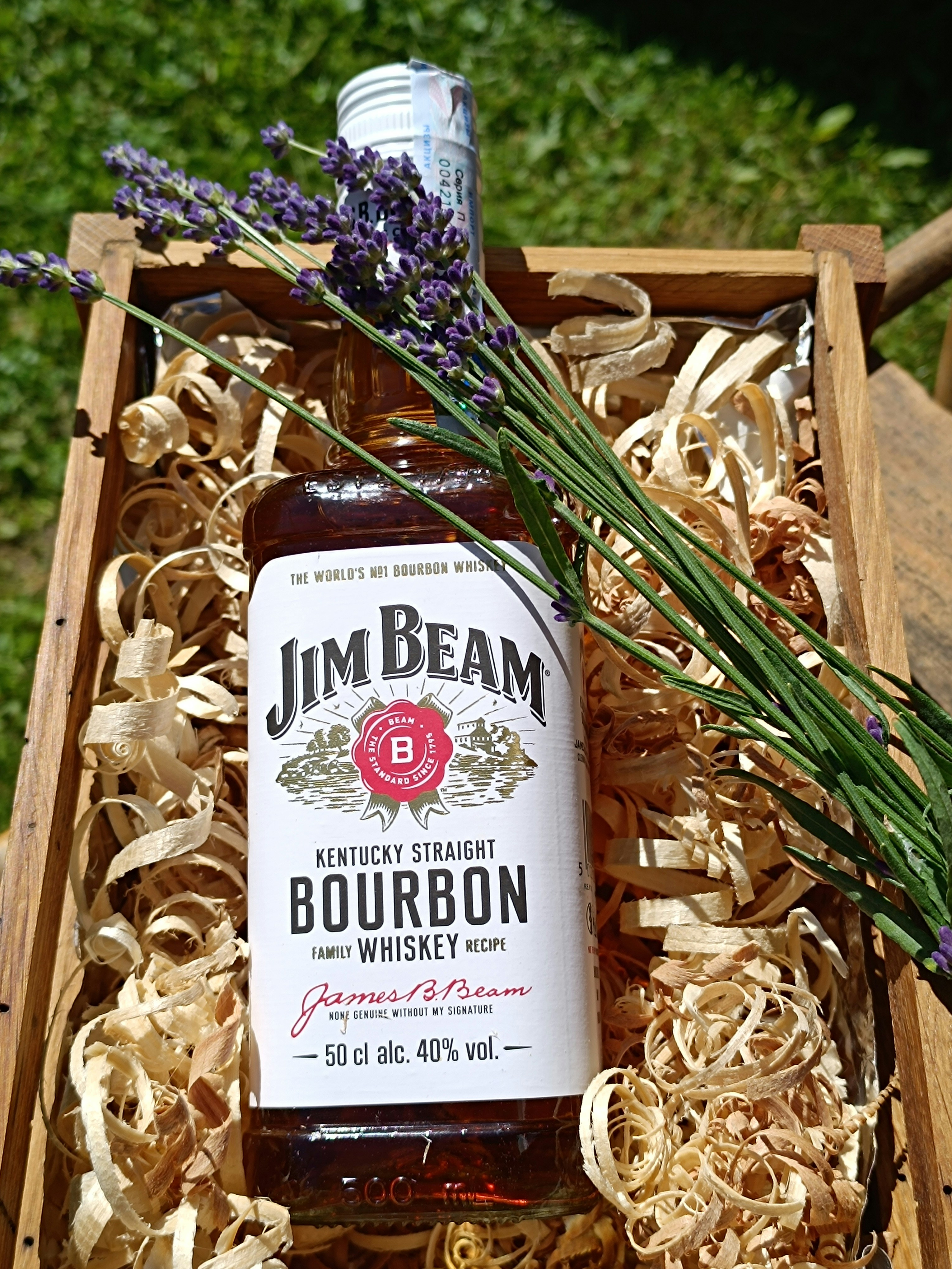 A bottle of Jim Beam bourbon nestled in wood shavings inside a wooden crate, with lavender sprigs laid across the label. A simple still-life showcasing packaging and garnish.