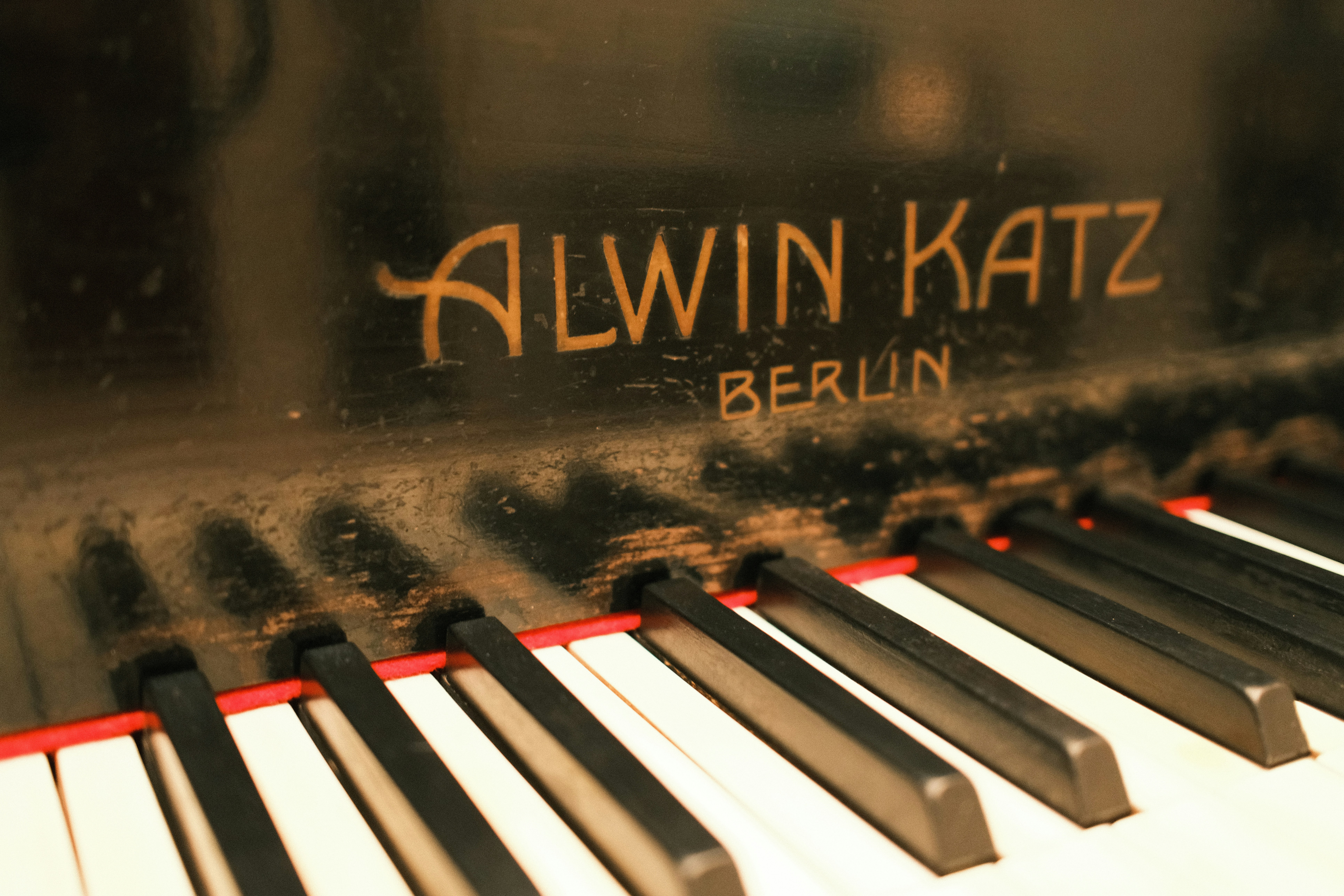 Close-up of an old Alwin Katz Berlin piano, focusing on the brand inscription above the keys with warm amber lighting and textured dust on the surface.