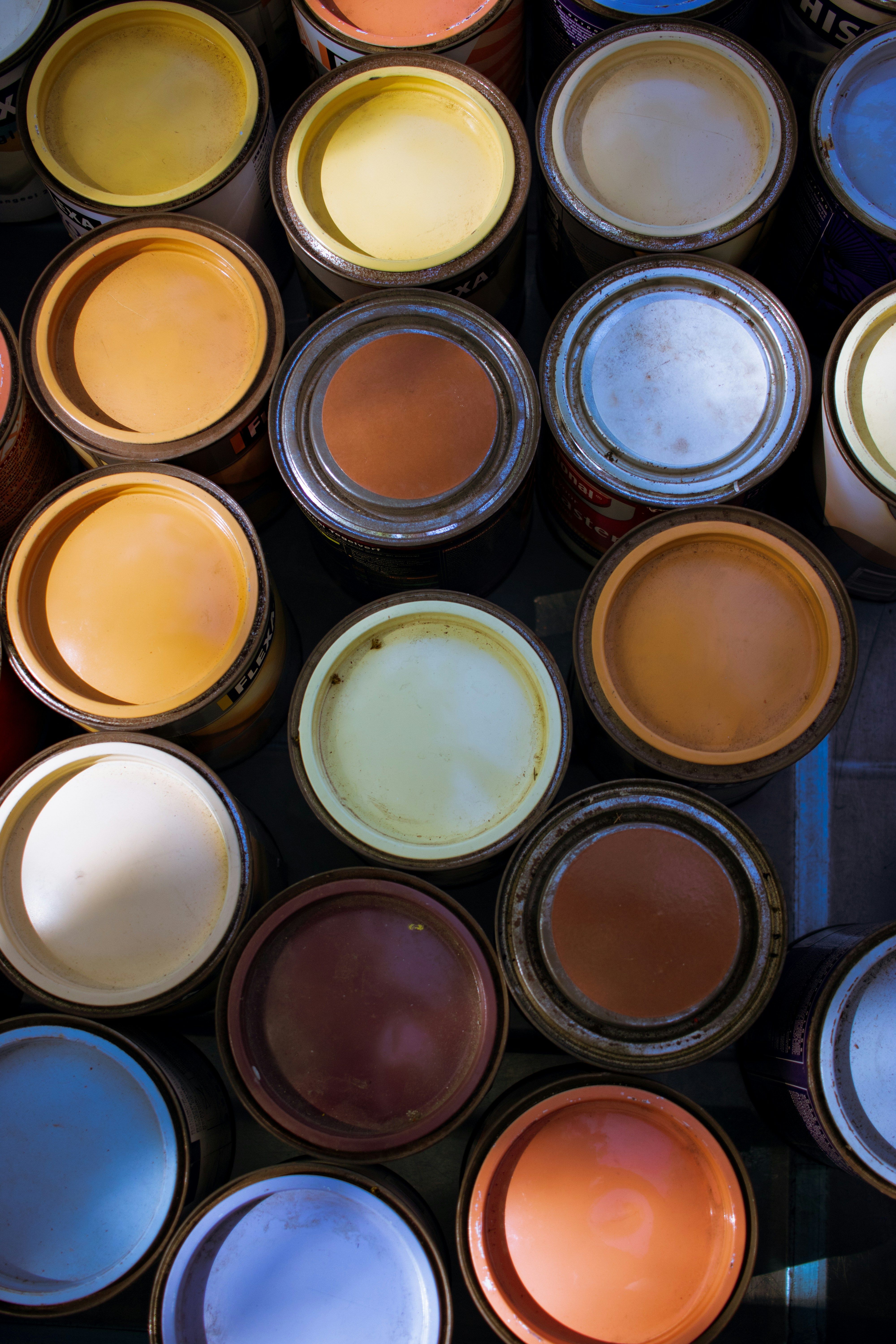 Close-up of open paint cans arranged in a dense grid, revealing a spectrum of dried and fresh colors; emphasizes color variety and texture of the paint surfaces.