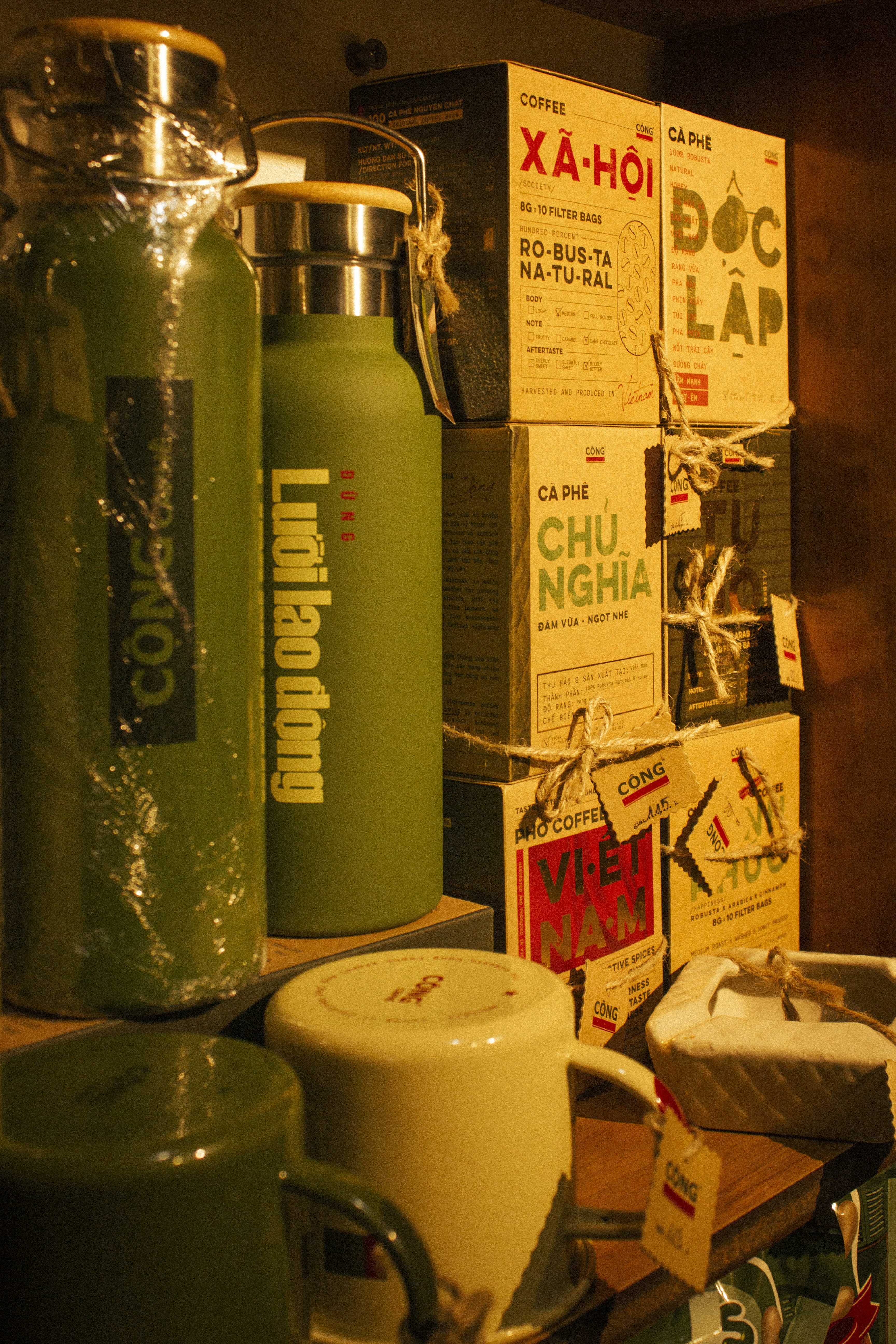 Warm-toned display of green water bottles and cardboard coffee boxes stacked on a shelf, with mugs in the foreground. A still-life arrangement emphasizing color and texture.
