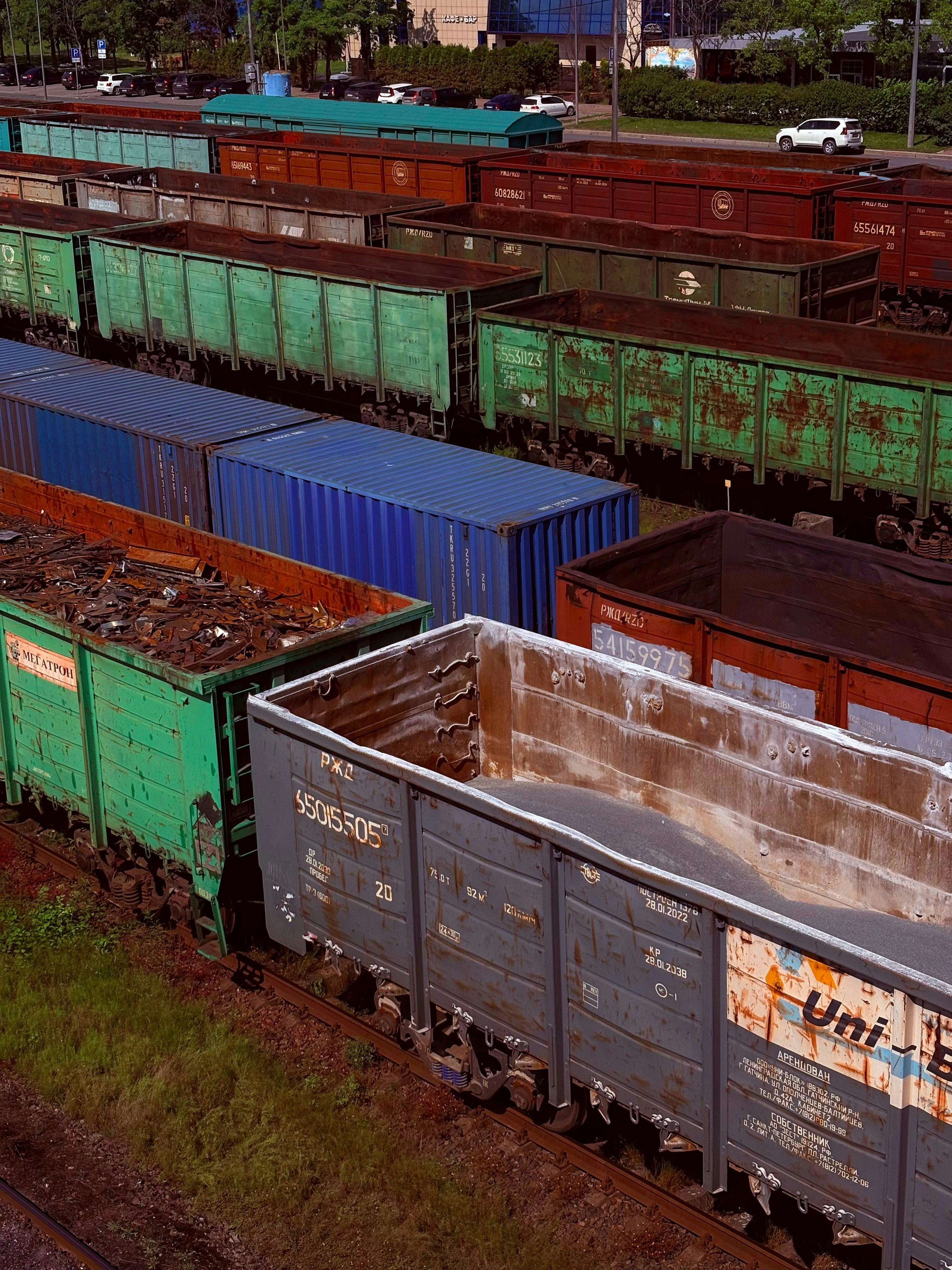 A hillside yard of weathered freight cars in green, blue, and rust hues stacked along rails; the scene emphasizes industrial texture and color variety.