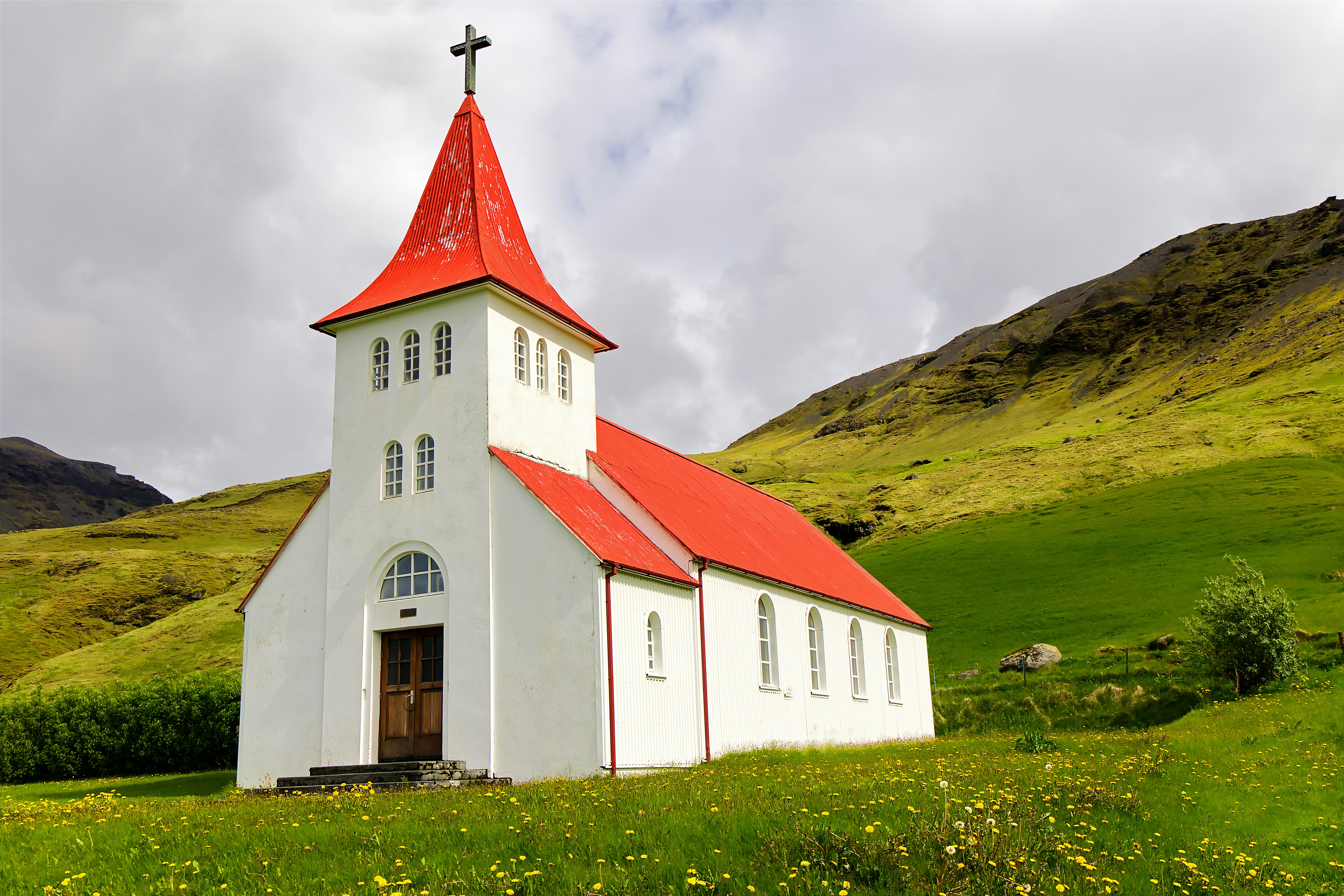 White church with red roof sits on a green Meadow under a cloudy sky; a small emblematic chapel in a pastoral landscape. Photograph.