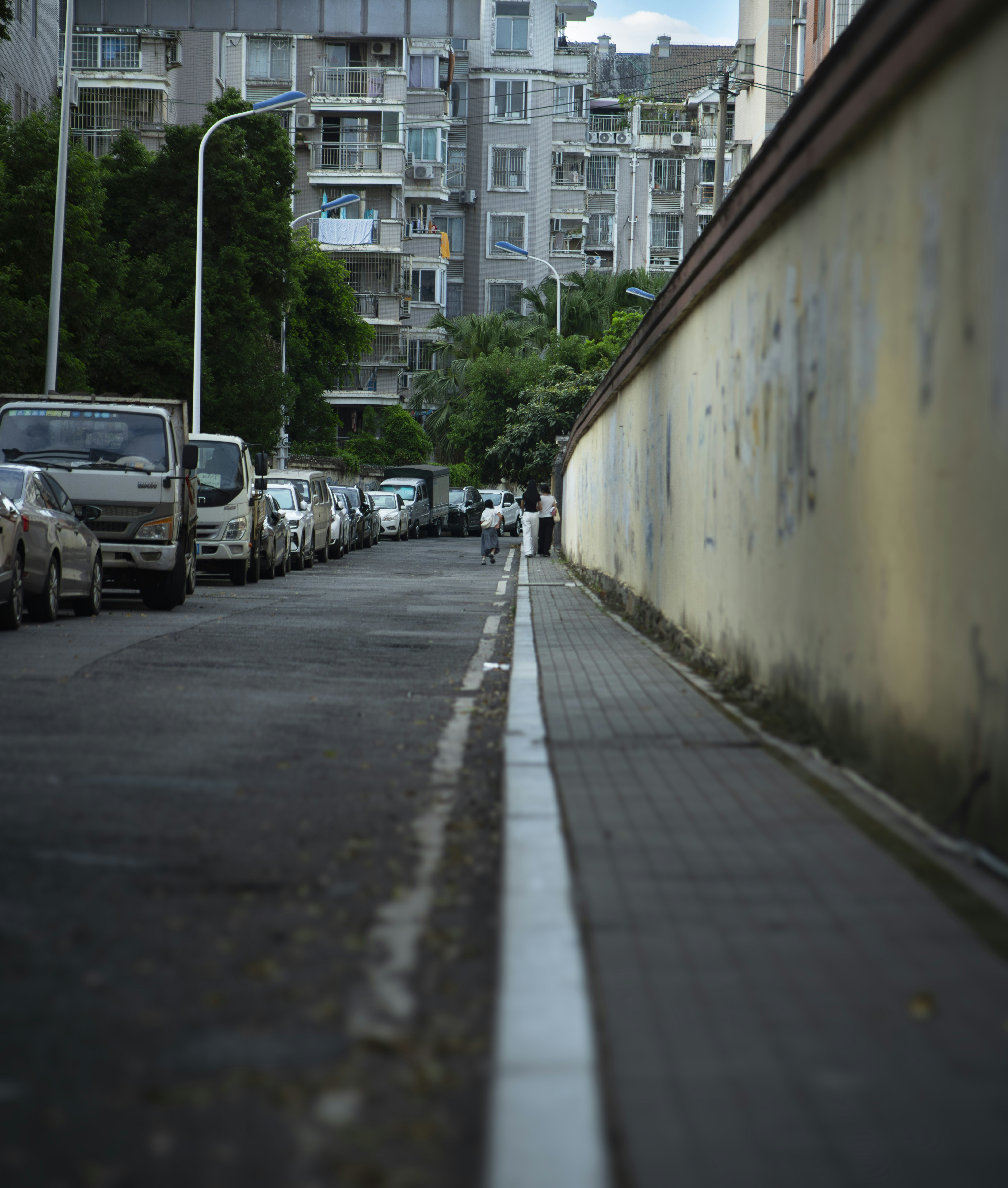 Narrow urban street lined with parked cars and a tall, weathered wall on the right, leading toward a distant group of pedestrians near mid-block; residential buildings rise in the background. (photograph)  
