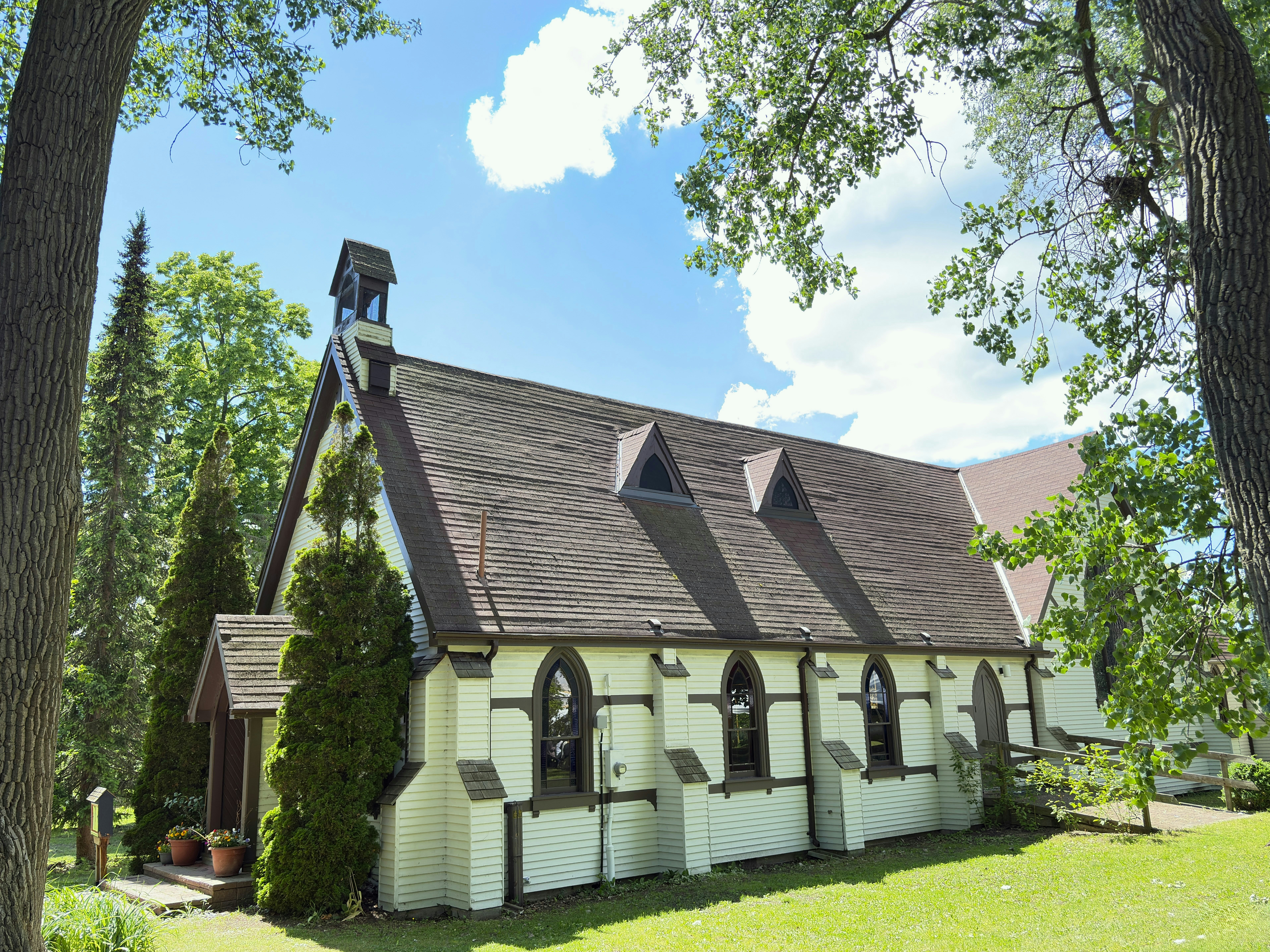 A quaint light-green wooden chapel with a steep roof sits among tall trees under a blue sky; a serene rural church scene with arched windows and trimmed lawn.