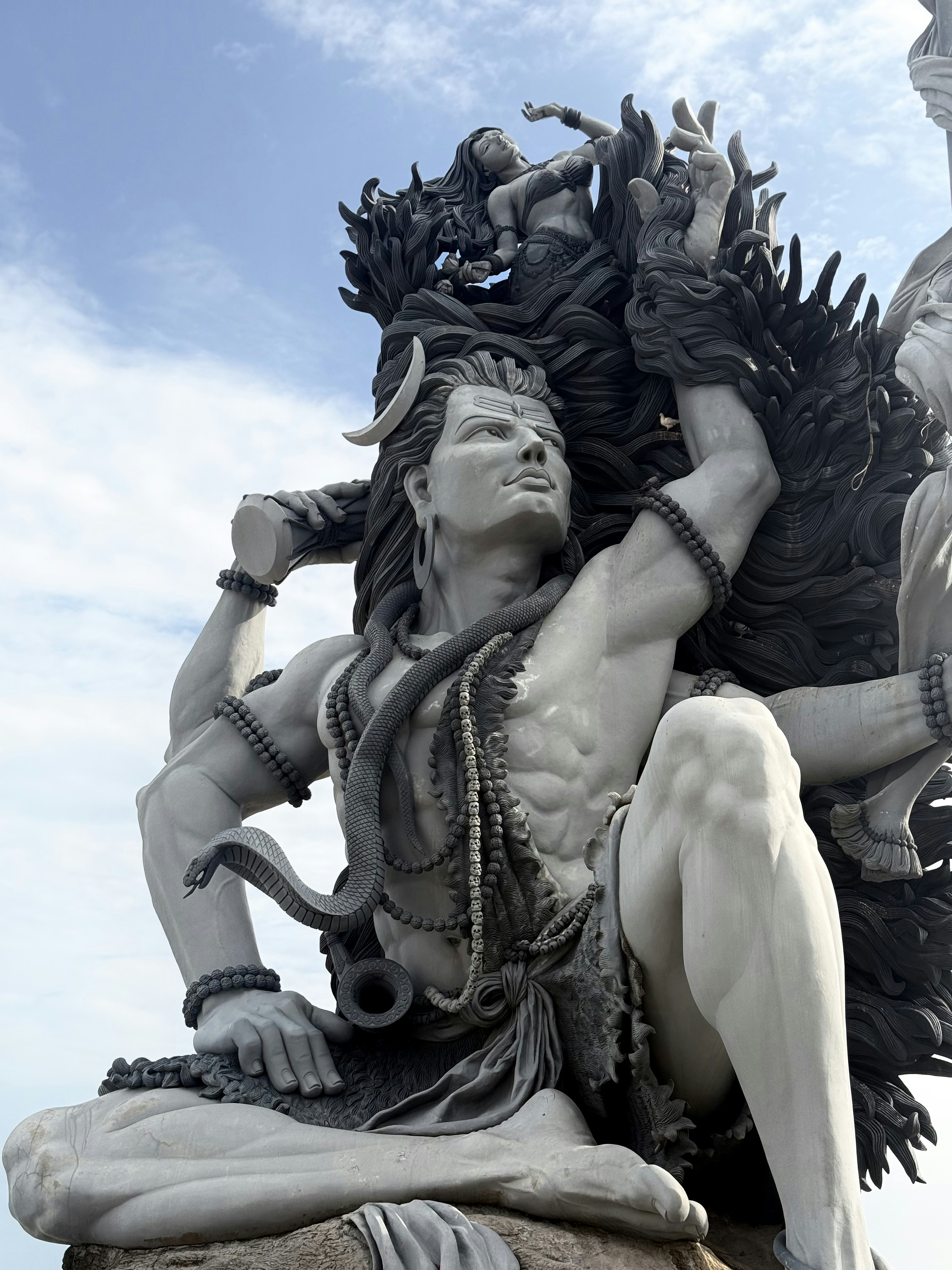 A monumental white marble statue of Lord Shiva captured from a dramatic low angle against an open sky, radiating raw divine power and meditative stillness simultaneously. The sculpture is rendered with extraordinary detail — the triple-lined tilaka on the forehead, a serpent coiled across the chest, rudraksha beads at the wrists, and wildly flowing jata locks from which a smaller dancing figure emerges above. Every muscle and adornment speaks to the fierce yet contemplative duality at the heart of Shaivite iconography.