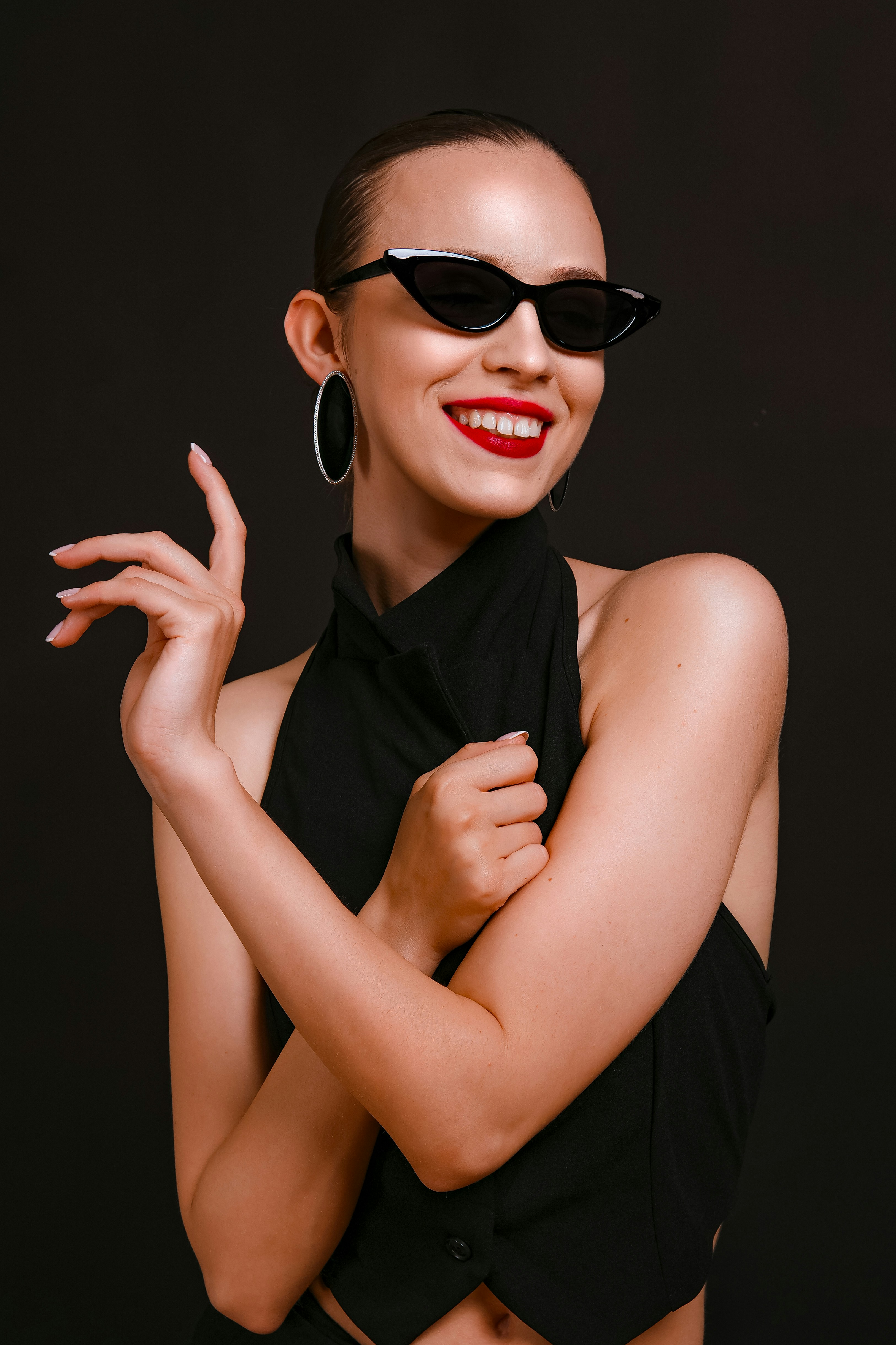 High-contrast portrait of a stylish woman wearing cat-eye sunglasses and red lipstick against a dark backdrop; the composition emphasizes her smile and confident pose. Logo-free studio shot.