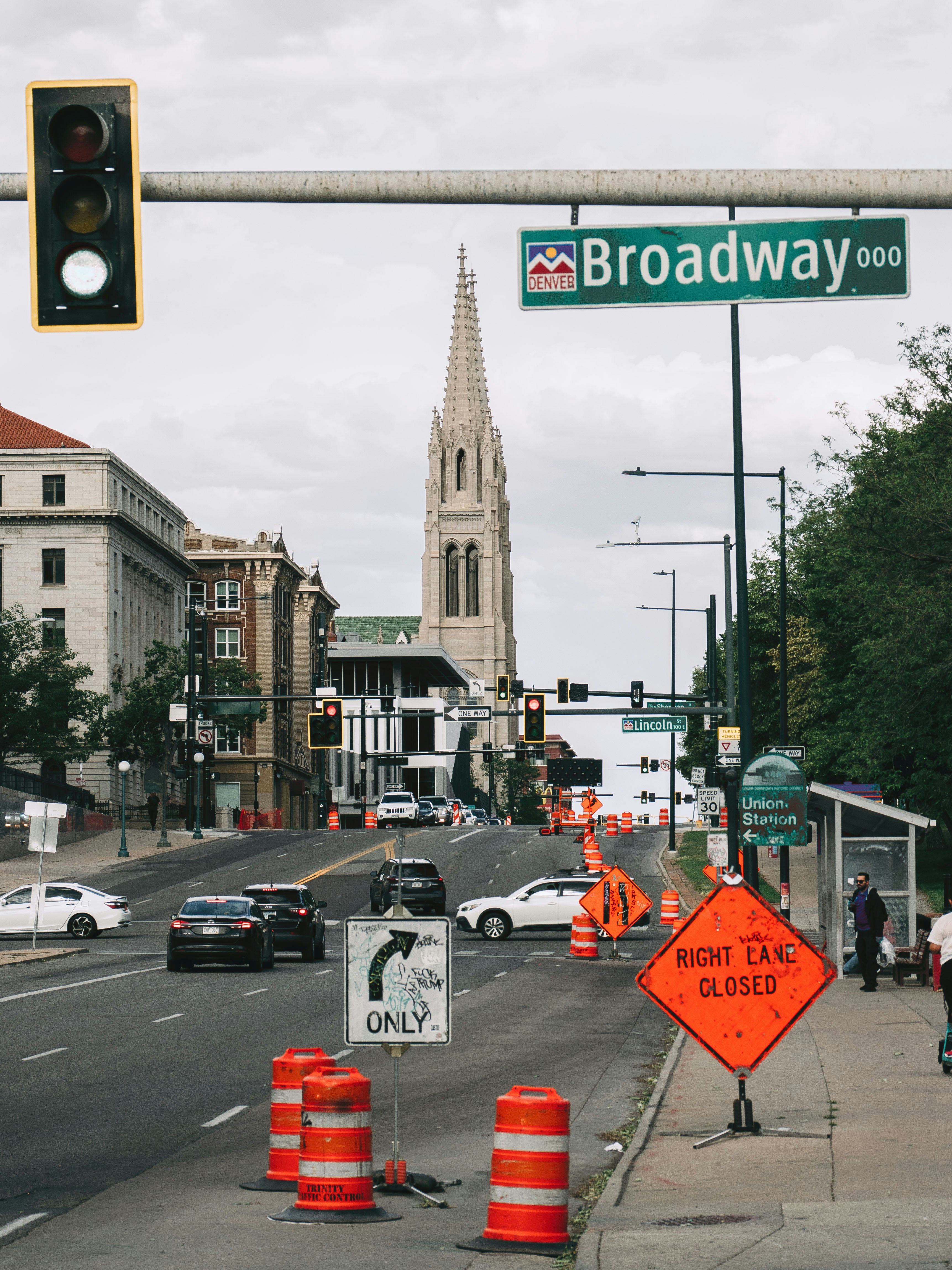A busy urban street with construction cones and a bright orange sign leads toward a distant cathedral spire; the scene captures city life and ongoing work in a downtown corridor. This is a photograph.