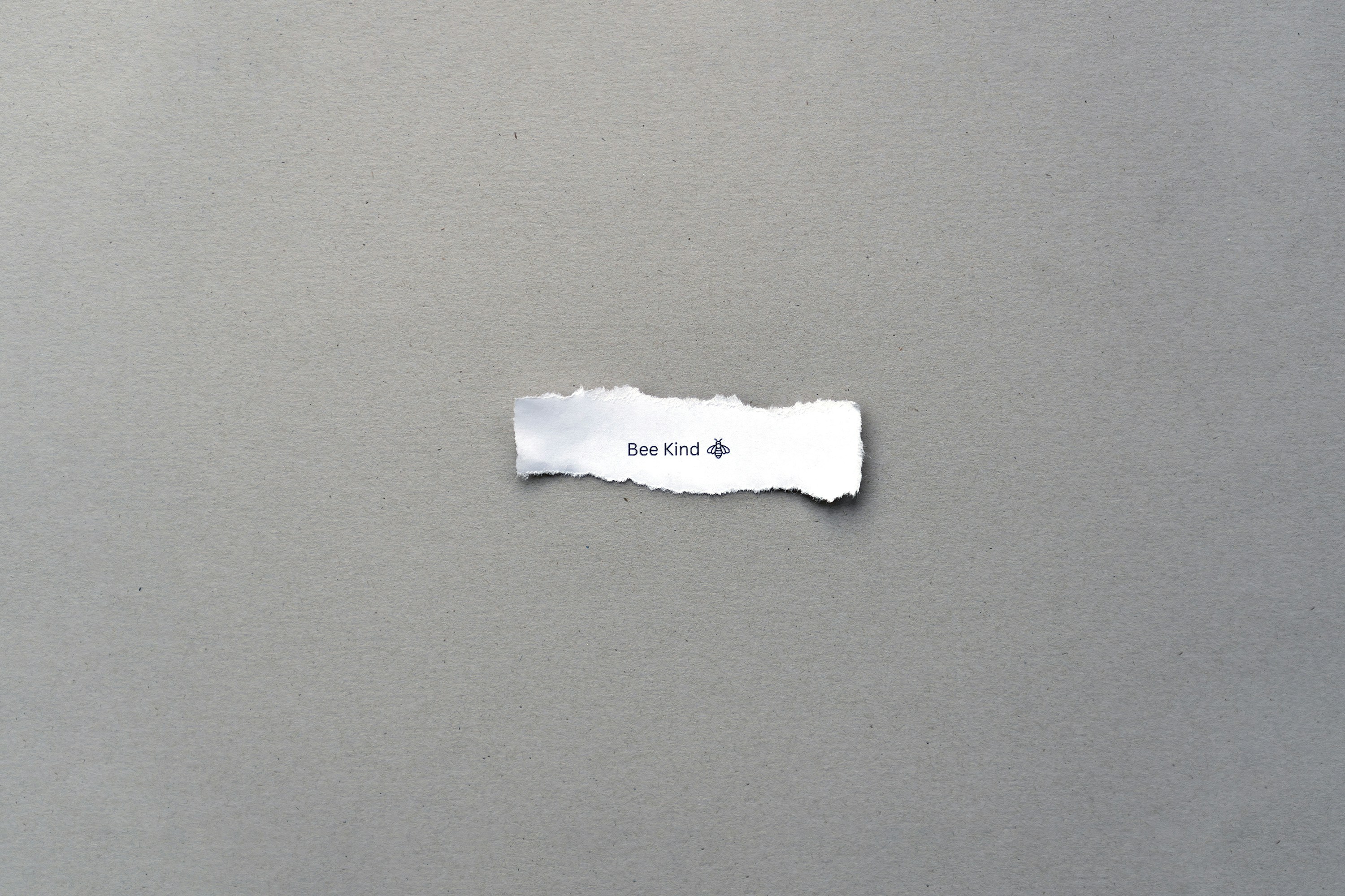 Small torn note reading 'Bee Kind' sits on a plain gray background; the piece acts as a minimal graphic element with subtle texture and shadow.
