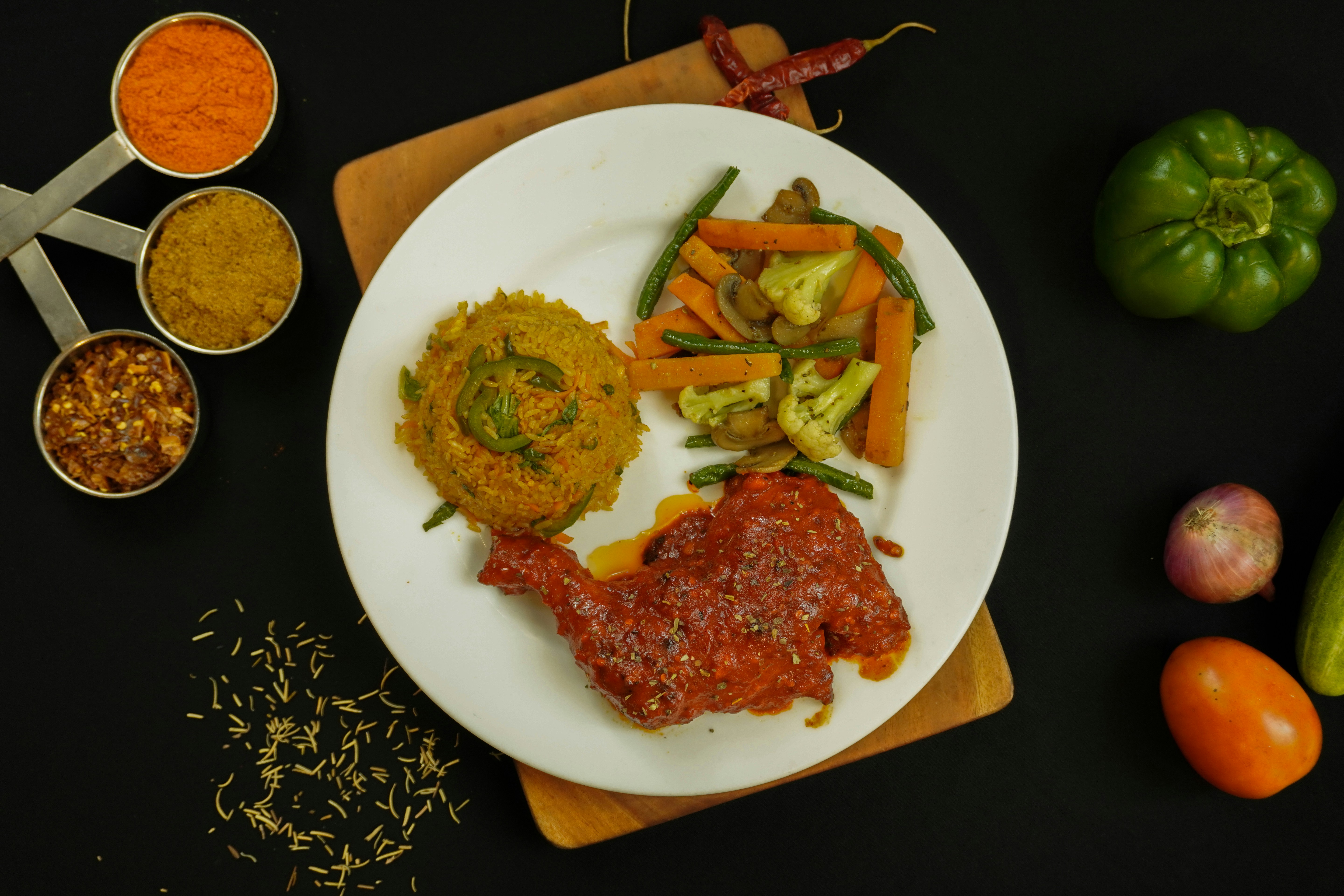 A plated meal featuring a curry-colored mound, roasted vegetables, and a red meat piece sits on a white plate against a black backdrop with fresh produce nearby. The scene is a still-life culinary setup and clearly shows a glossy sauce and garnish highlights.