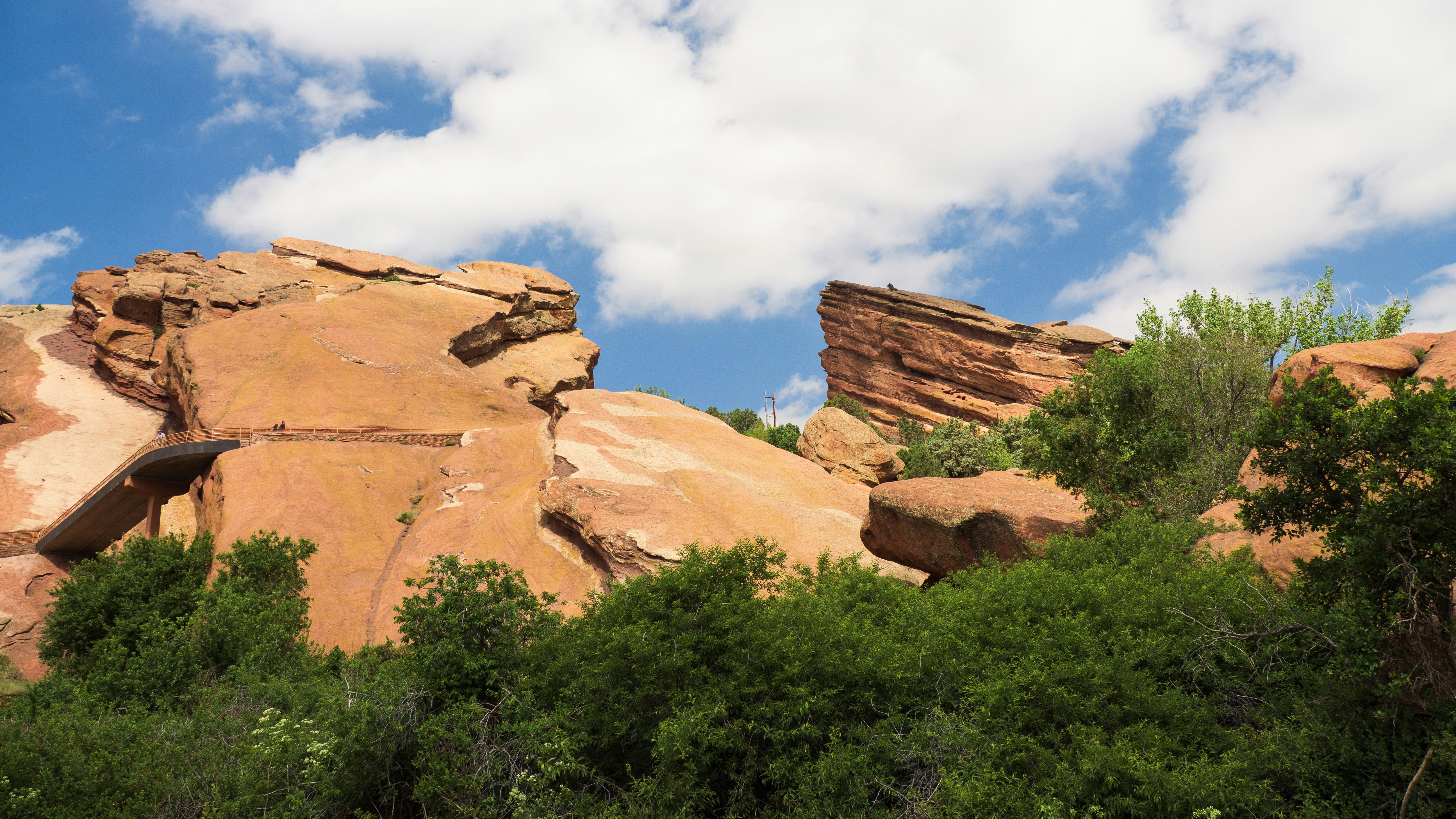 Sunlit red rock formations rise above dense green shrubs in a bright, clear landscape; the scene emphasizes geology and scale with a vivid blue sky.