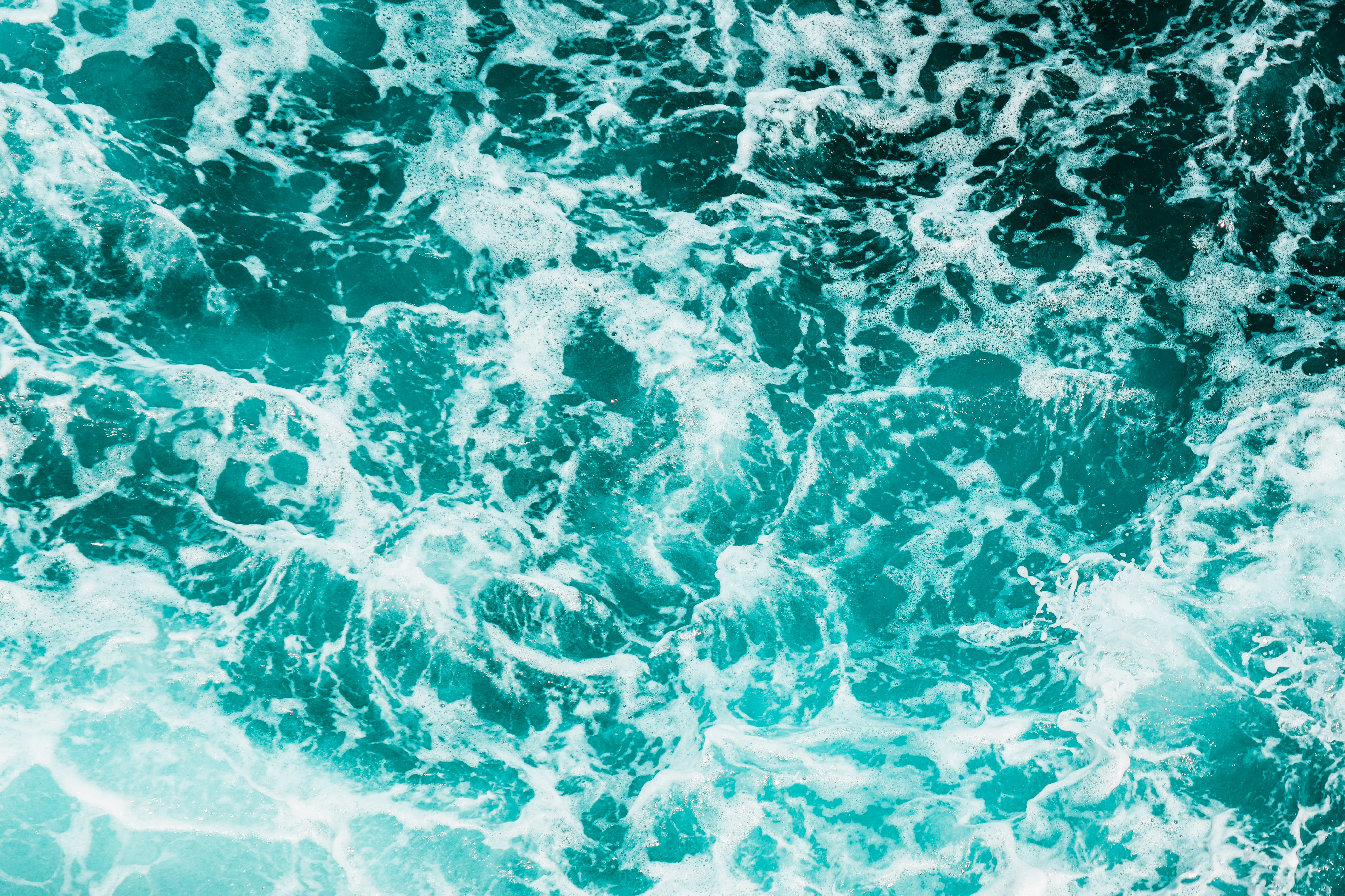 Vibrant turquoise waves churn along the shoreline, captured as a close-up abstraction of sea foam and surf textures. A dynamic, water-themed composition suitable for branding or background use.