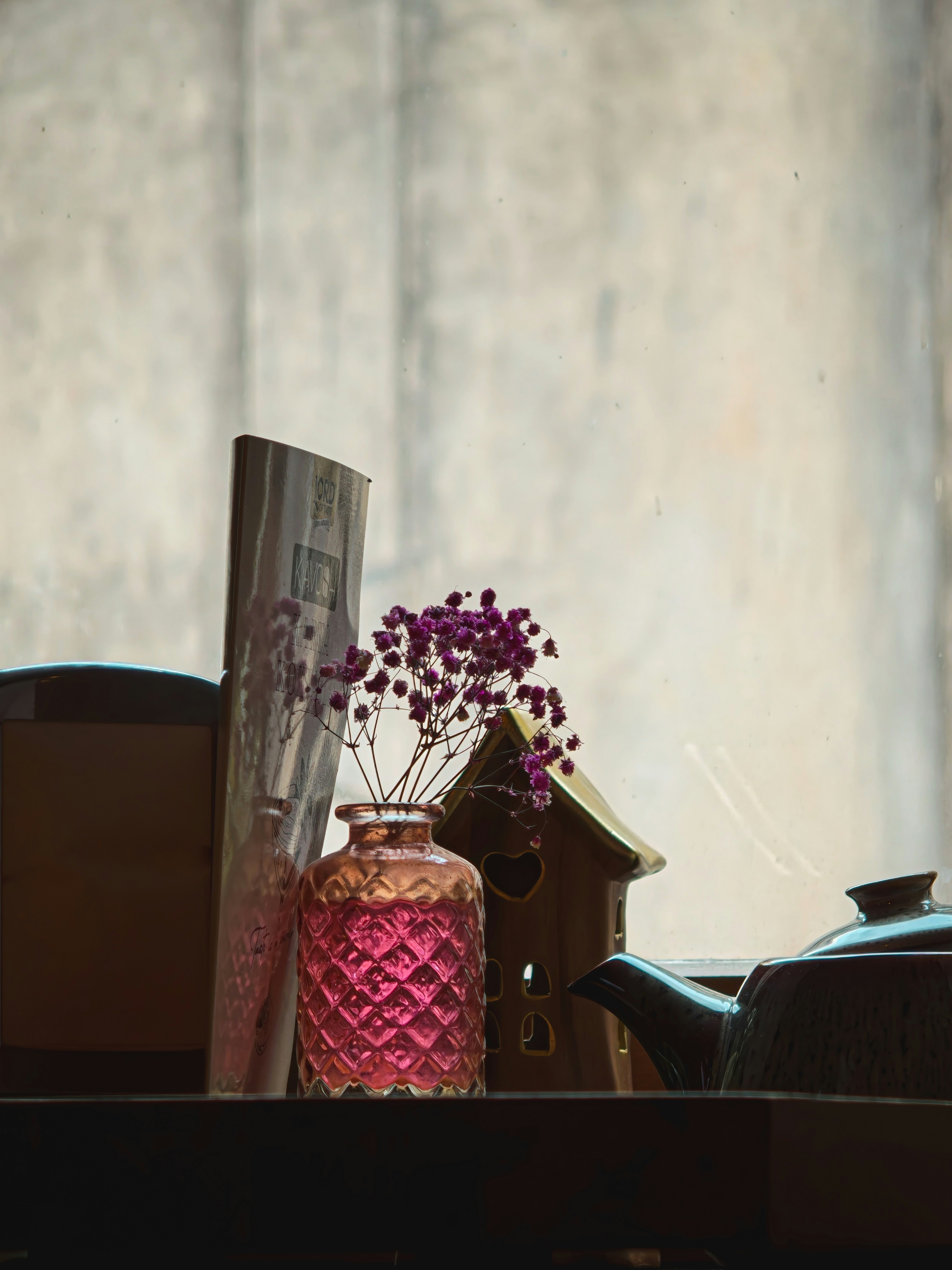 Purple sprigs in a pink glass vase anchor a still life on a sunlit shelf