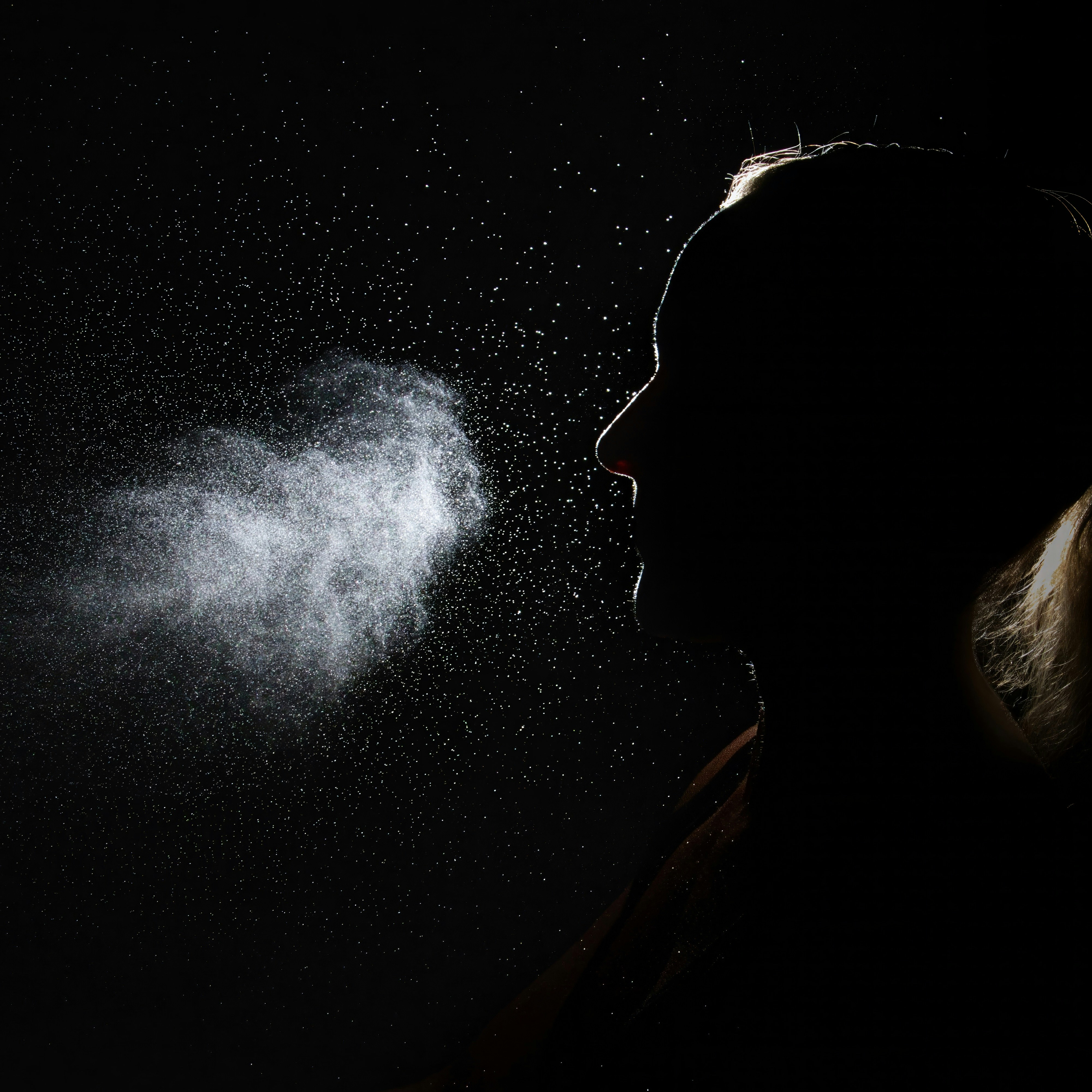 Silhouette portrait of a person exhaling a cloud of powder against a dark background, highlighting the breath and dust in motion. This is a photographic study focusing on negative space and texture.