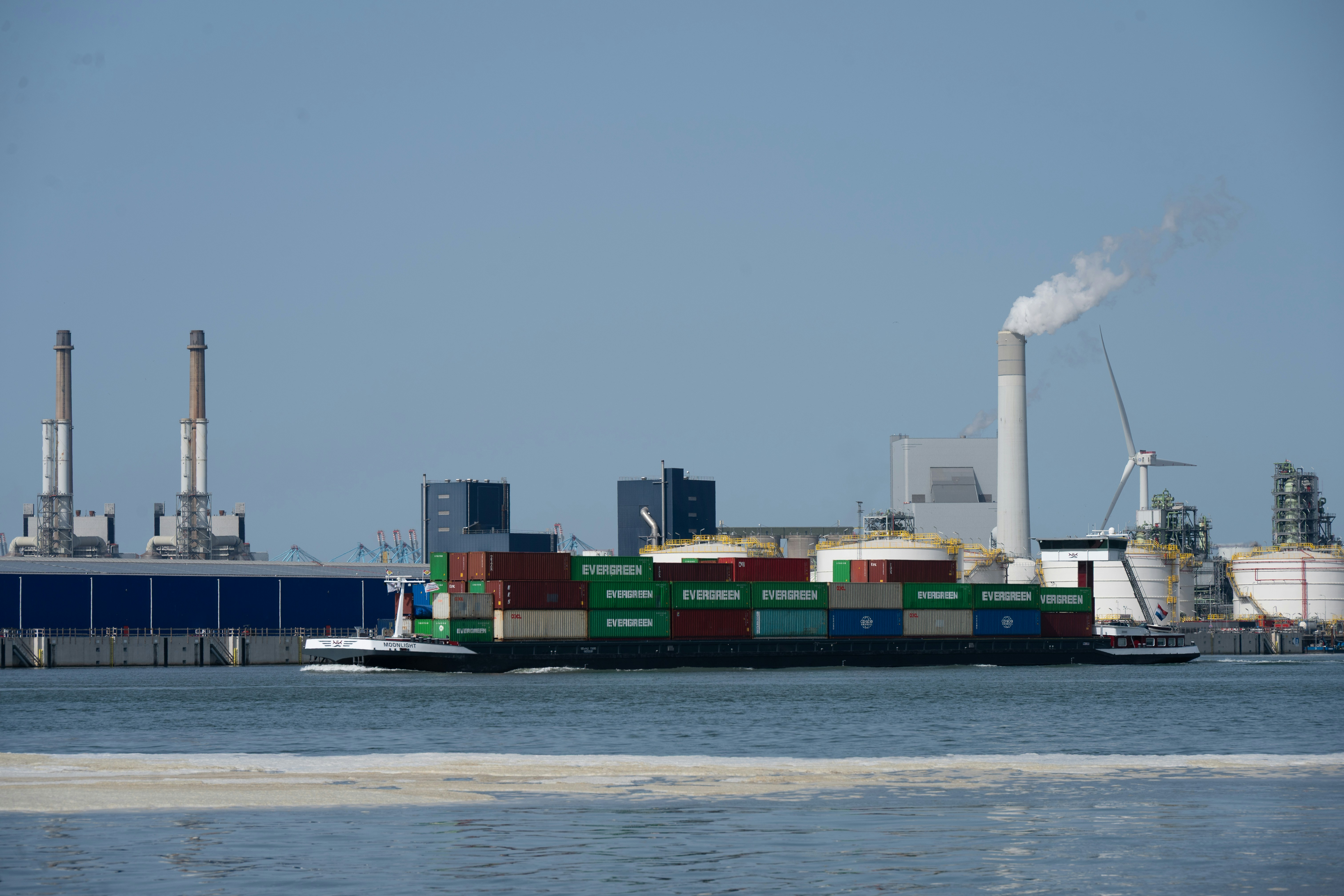 Container Ship gliding past an industrial shoreline under a calm sky