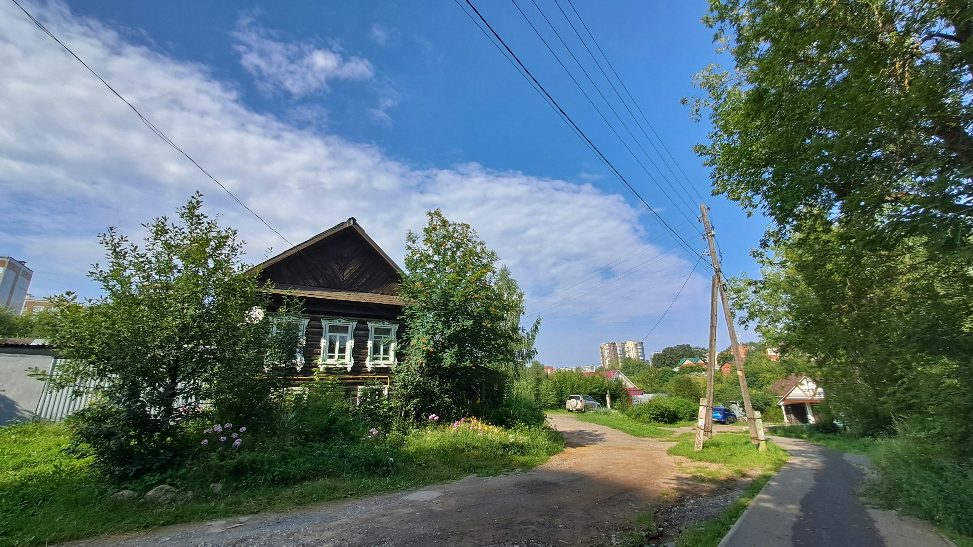Quiet rural path with an old wooden house on the left, overgrown garden, and a dirt road leading into a small village under a bright blue sky.