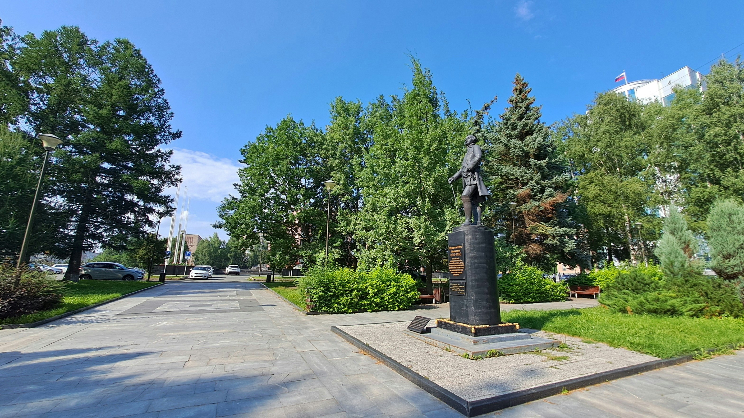 Sunlit park walkway with a prominent statue on a pedestal and surrounding greenery, capturing a calm civic space.