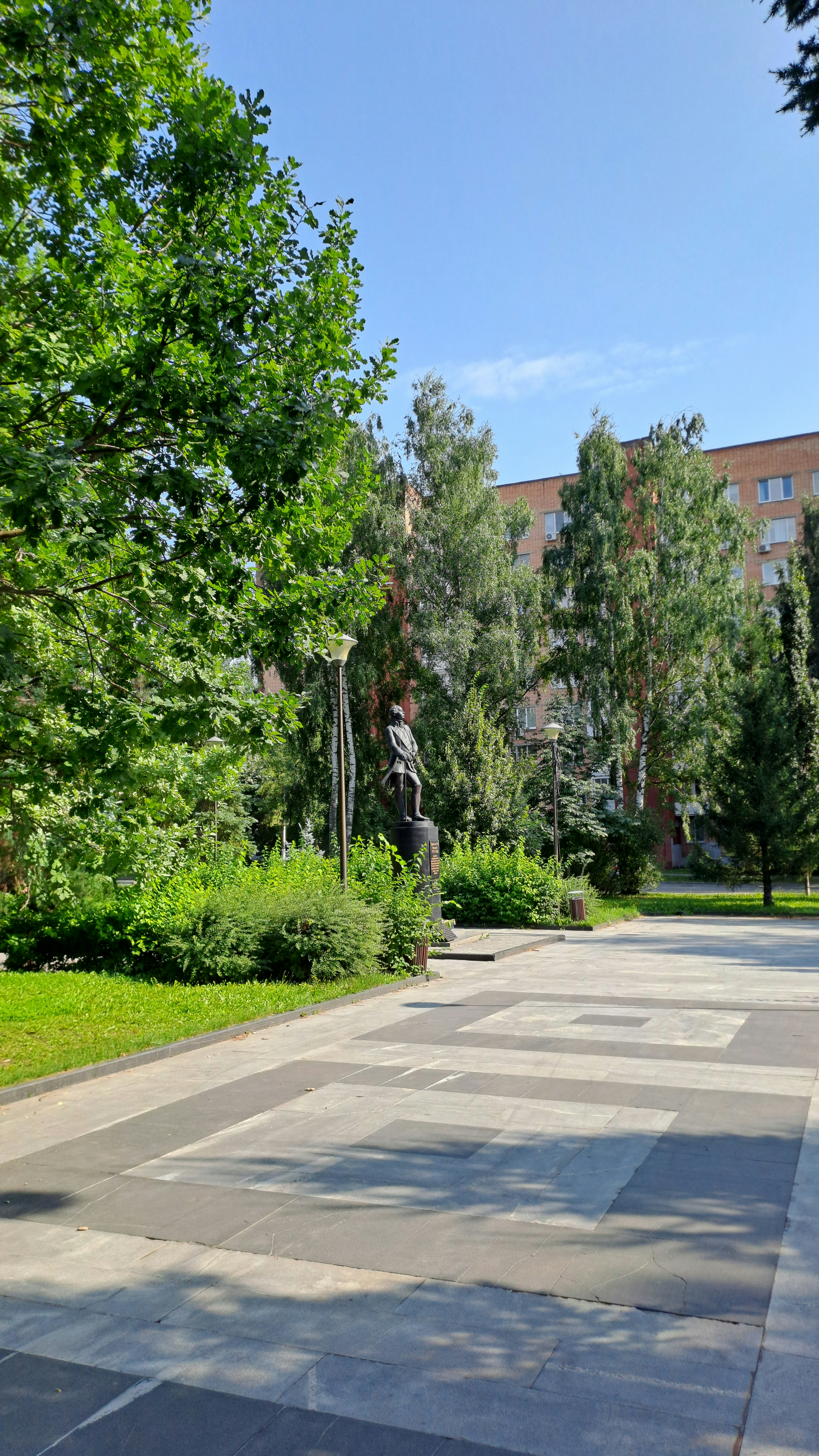 A sunlit urban park path leads to a statue surrounded by lush greenery and tall trees, with a building visible in the background.
