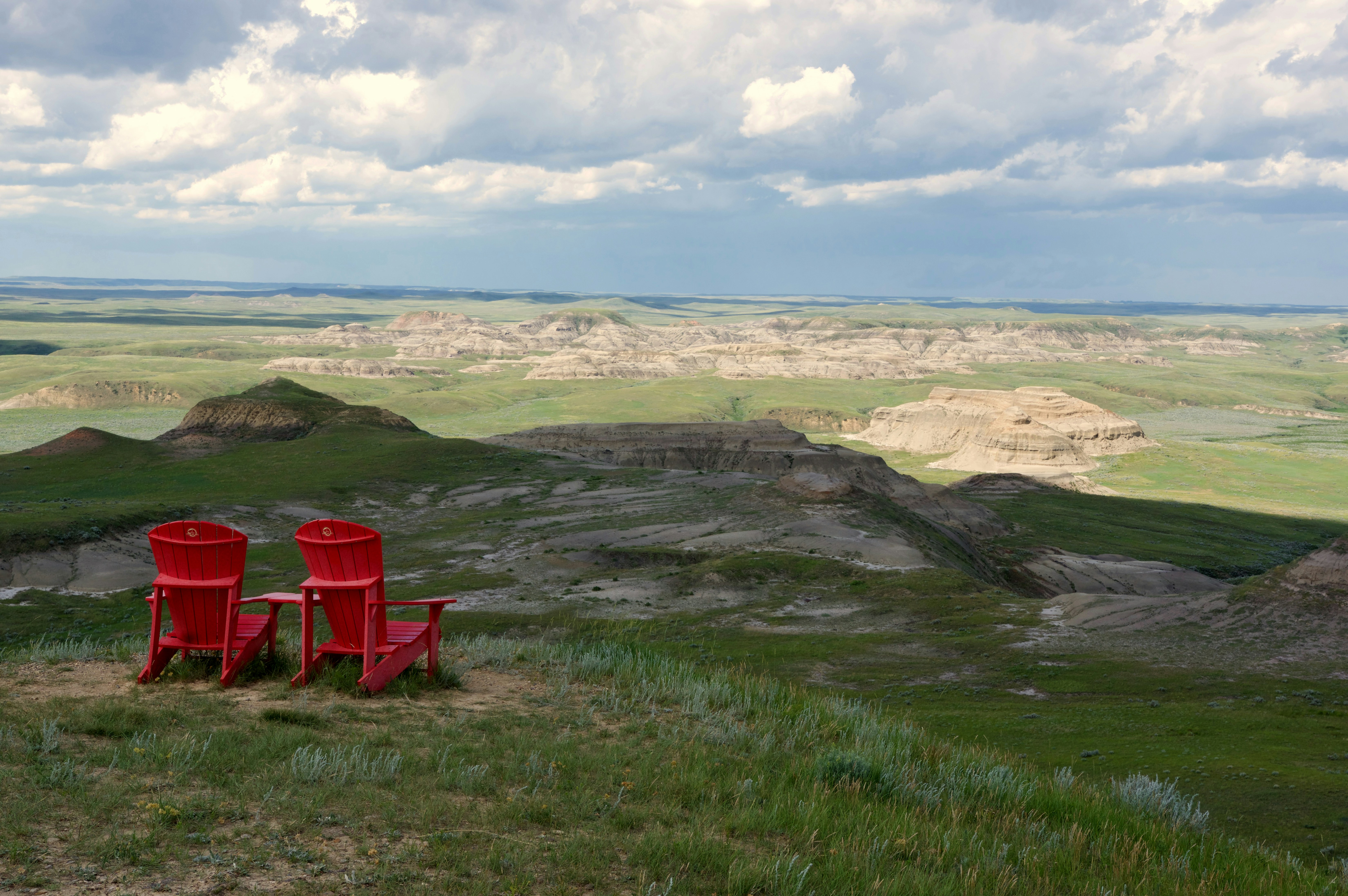 Two red Adirondack chairs sit on grassy foreground overlooking a vast eroded plateau and hills under a partly cloudy sky.