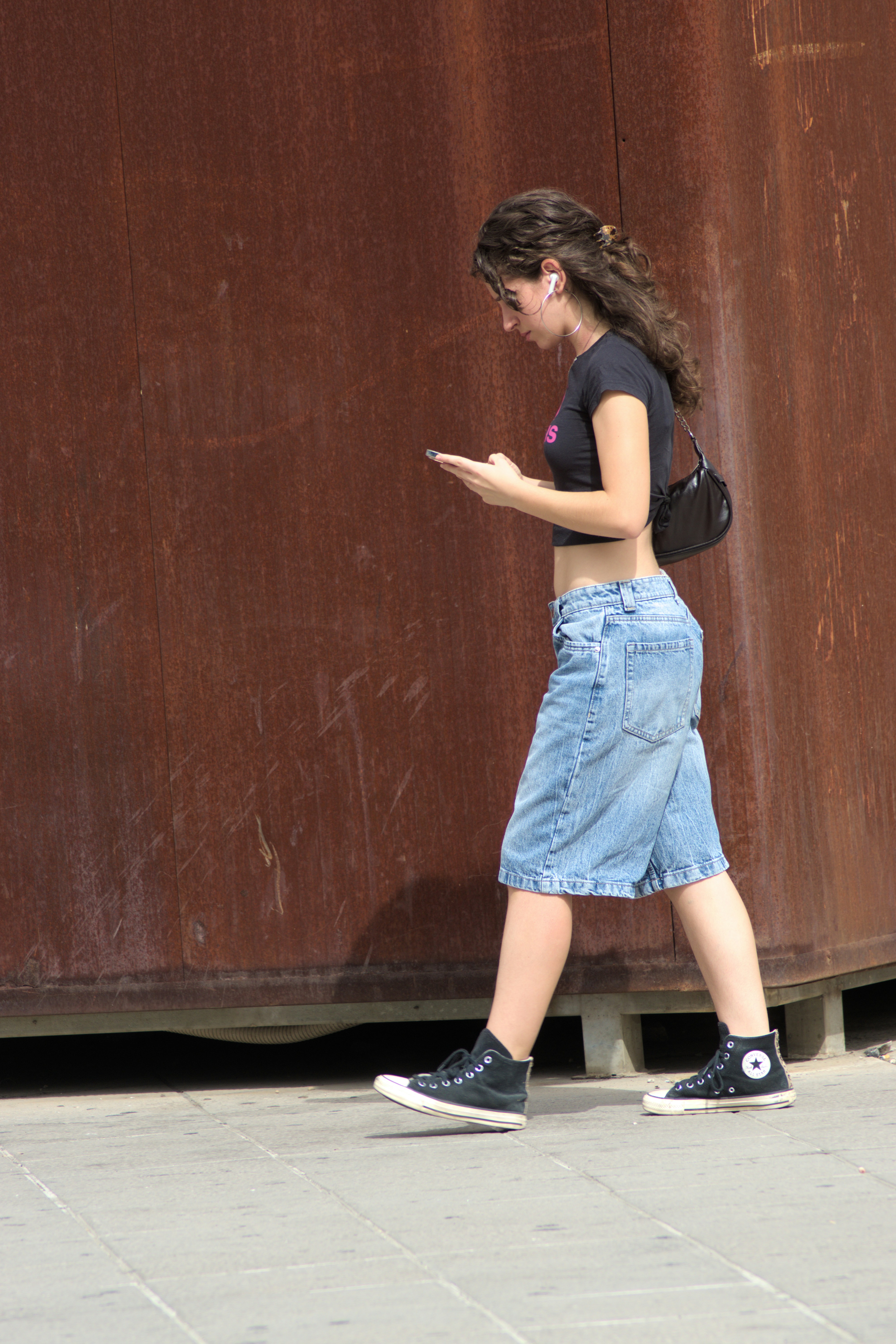 A young lady walking in the street with cellular and earphones. She seems to be in a hurry