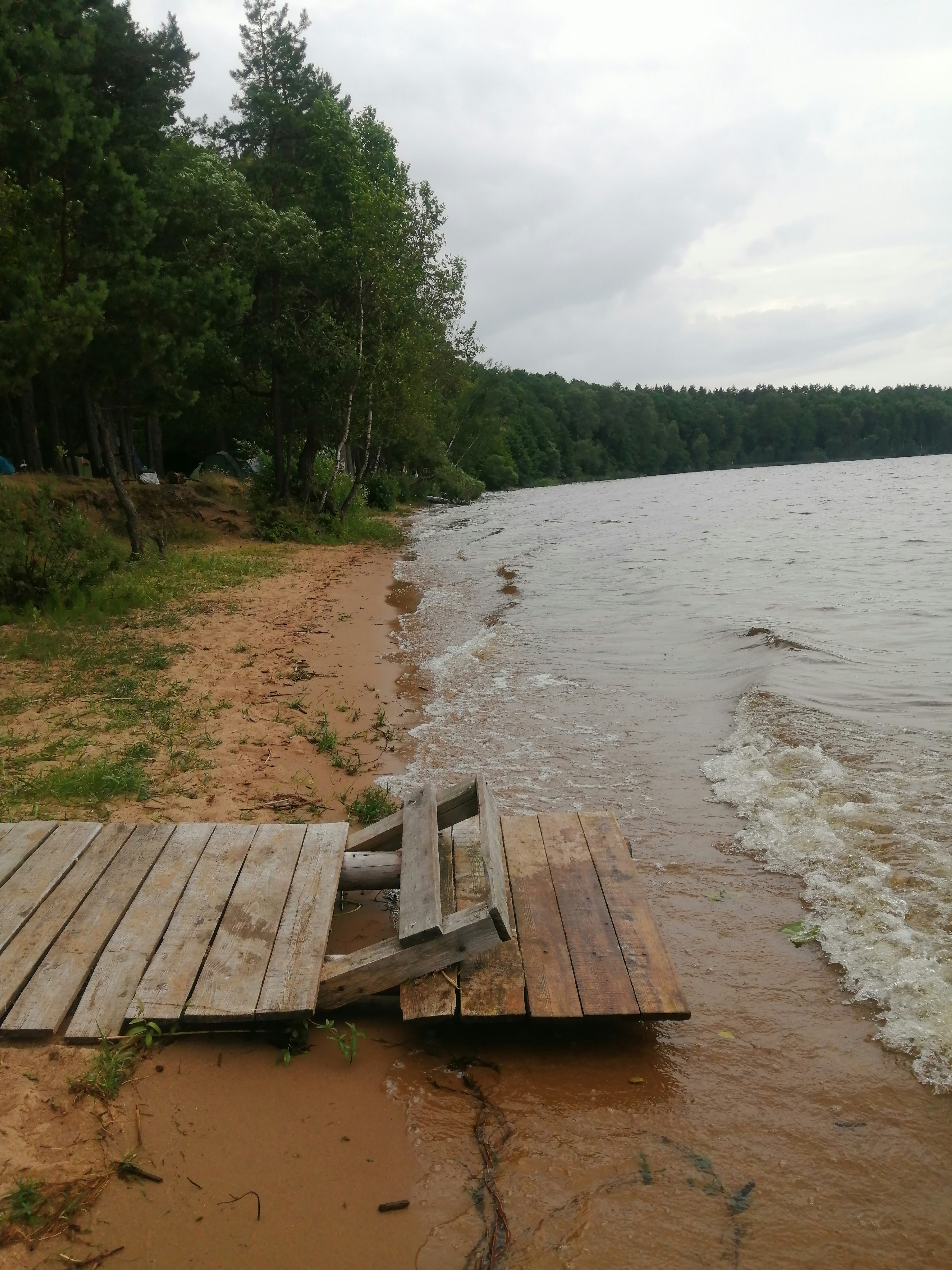 Wooden dock jutting into a calm lake along a sandy shoreline with a forest backdrop; a weathered structure rests at the water’s edge. (Note: this is a landscape scene; no logos.)