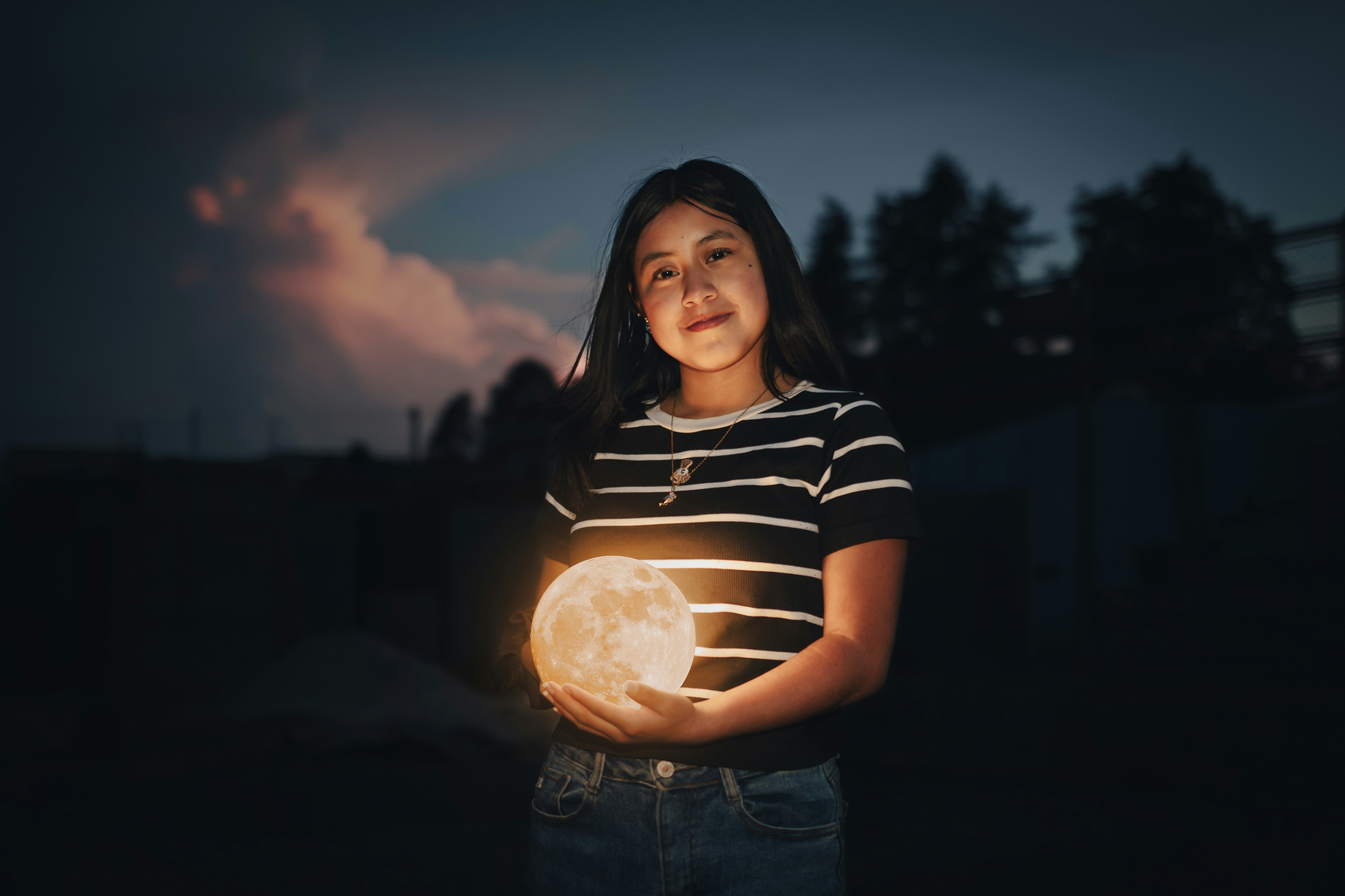 A young woman stands outdoors at dusk, cradling a softly glowing orb that lights her face. The scene emphasizes the contrast between the warm orb and the dark background, with the subject centered and foreground illumination highlighting her expression.