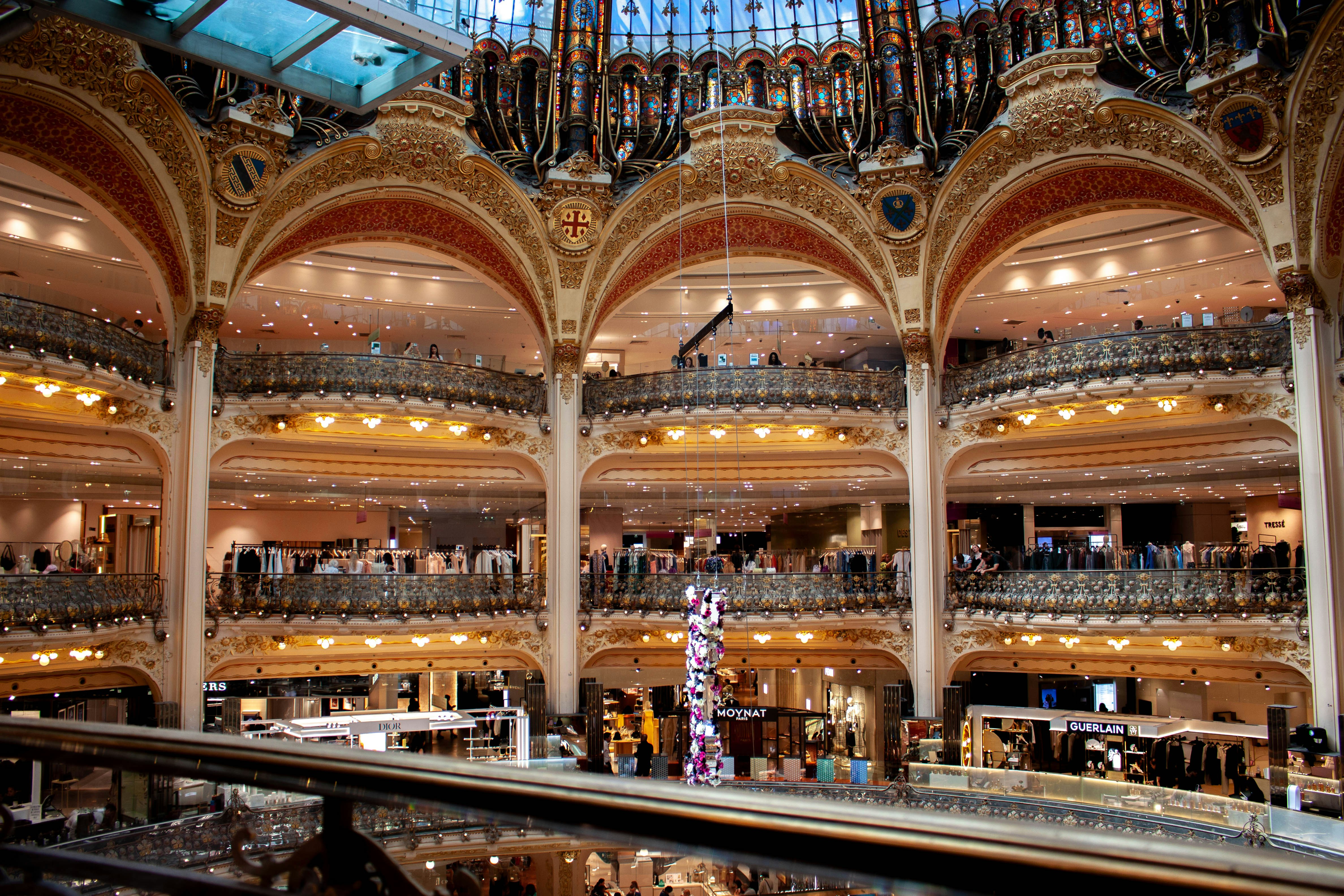 Ornate shopping atrium with gold-accent arches, balconies and shoppers around a central vertical display, a storefront-lined interior under a chandelier-lit ceiling.
