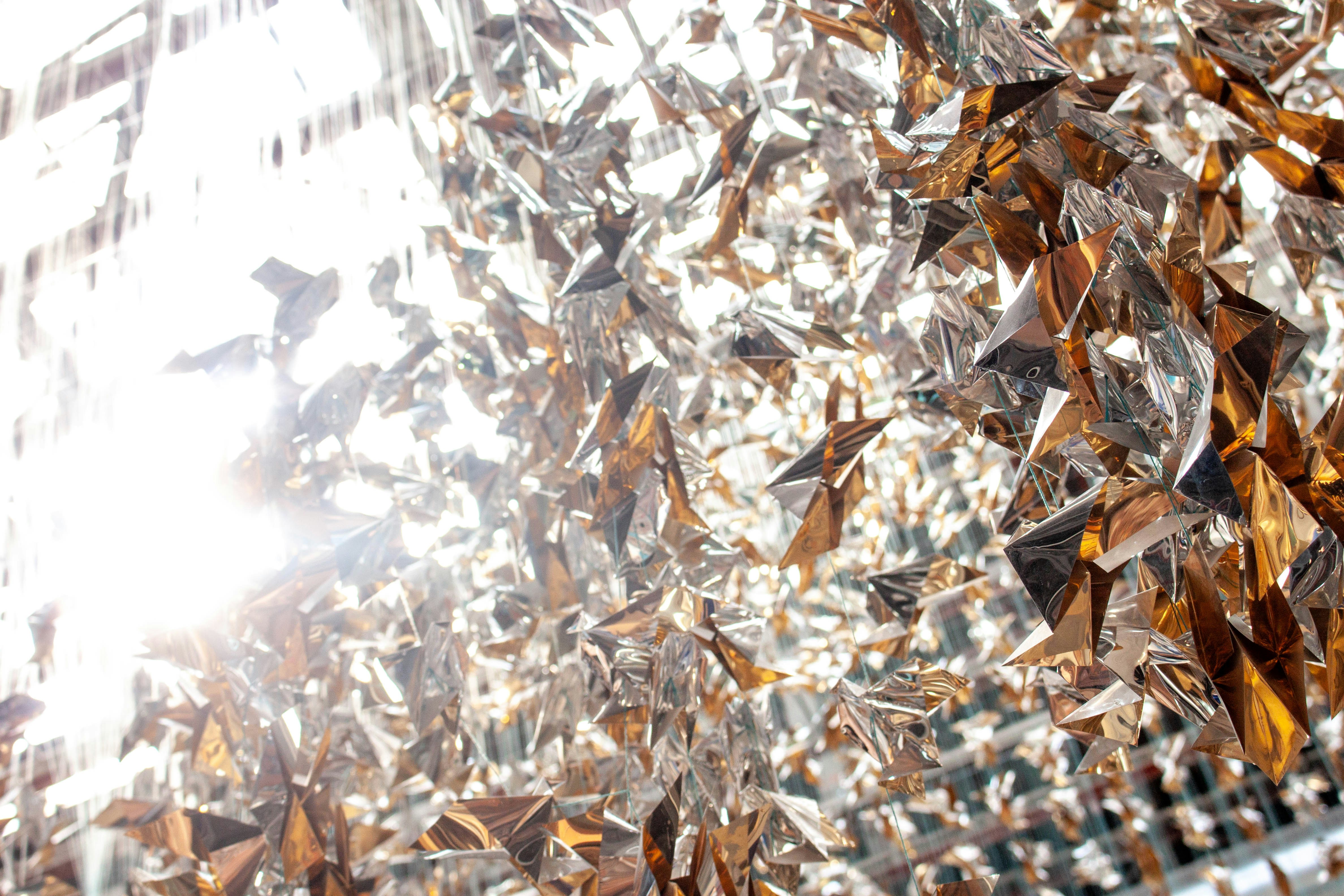 Close-up of metallic, copper-toned triangular leaves lit by bright sunlight, forming a reflective abstract texture; appears to be an installation or sculpture with a glassy, mirrored backdrop.