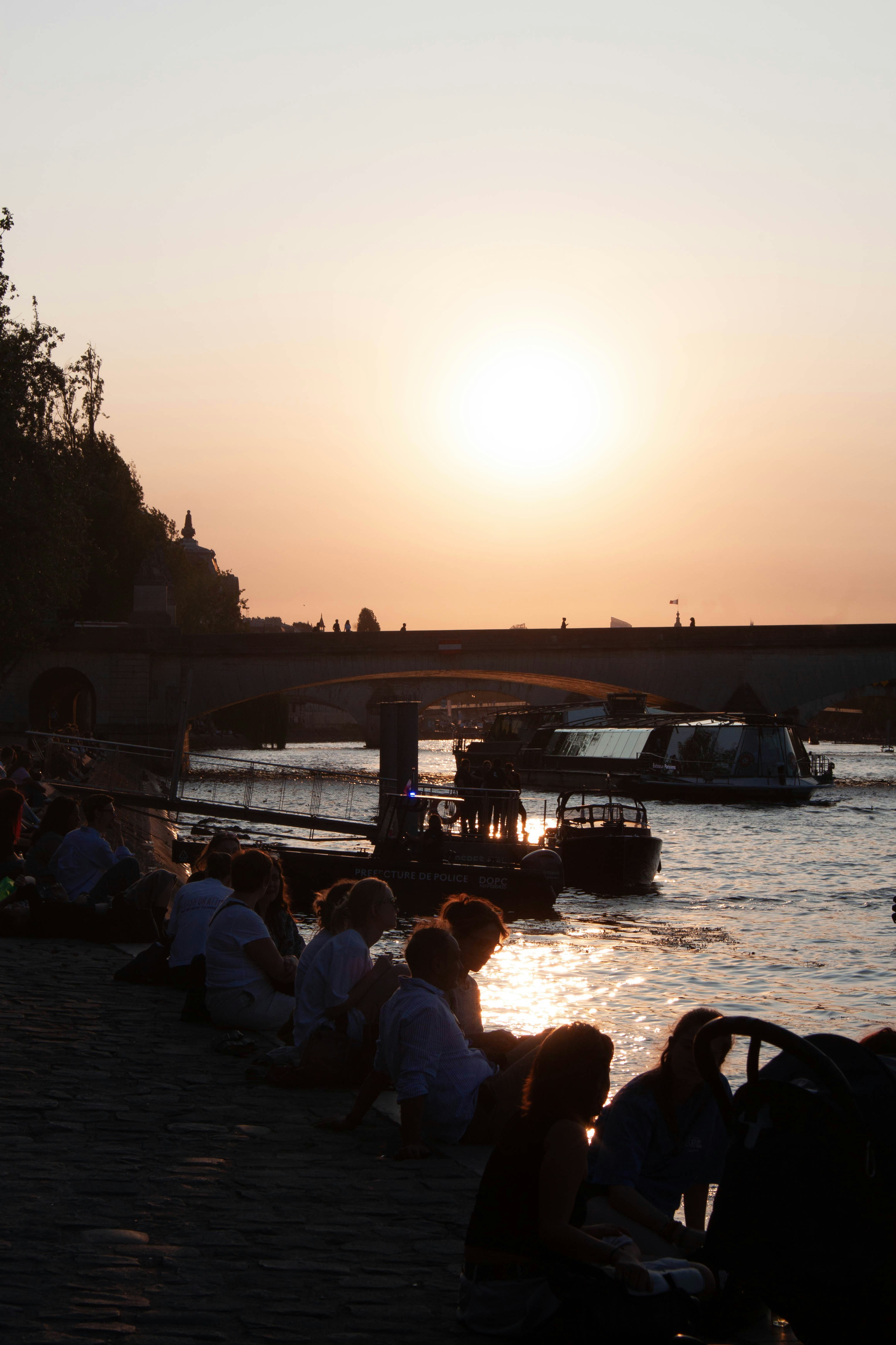 Sunset by the Riverbank: People, Boats and a Silhouetted Bridge