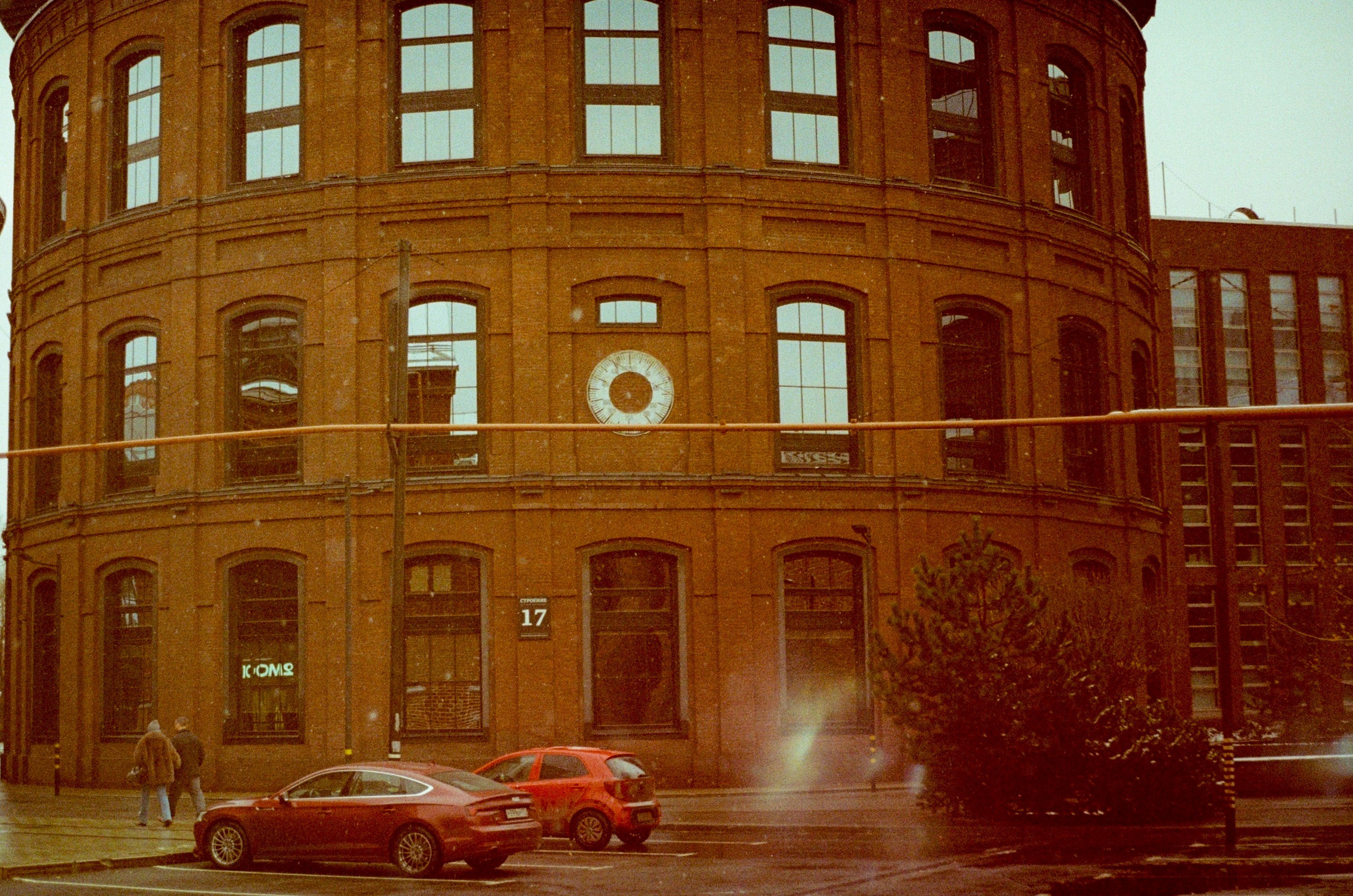 A slightly out‑of‑focus film photograph of a brick building of the former Moscow Gas Works, with a parking lot and cars visible in the foreground. Shot on analog film in November, the image carries a moody, nostalgic industrial atmosphere with soft grain and muted autumn tones.