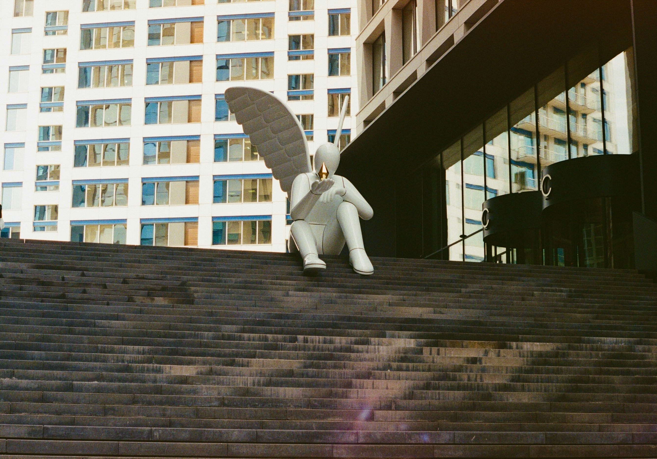 White angel statue sitting on stairs outside a modern building.