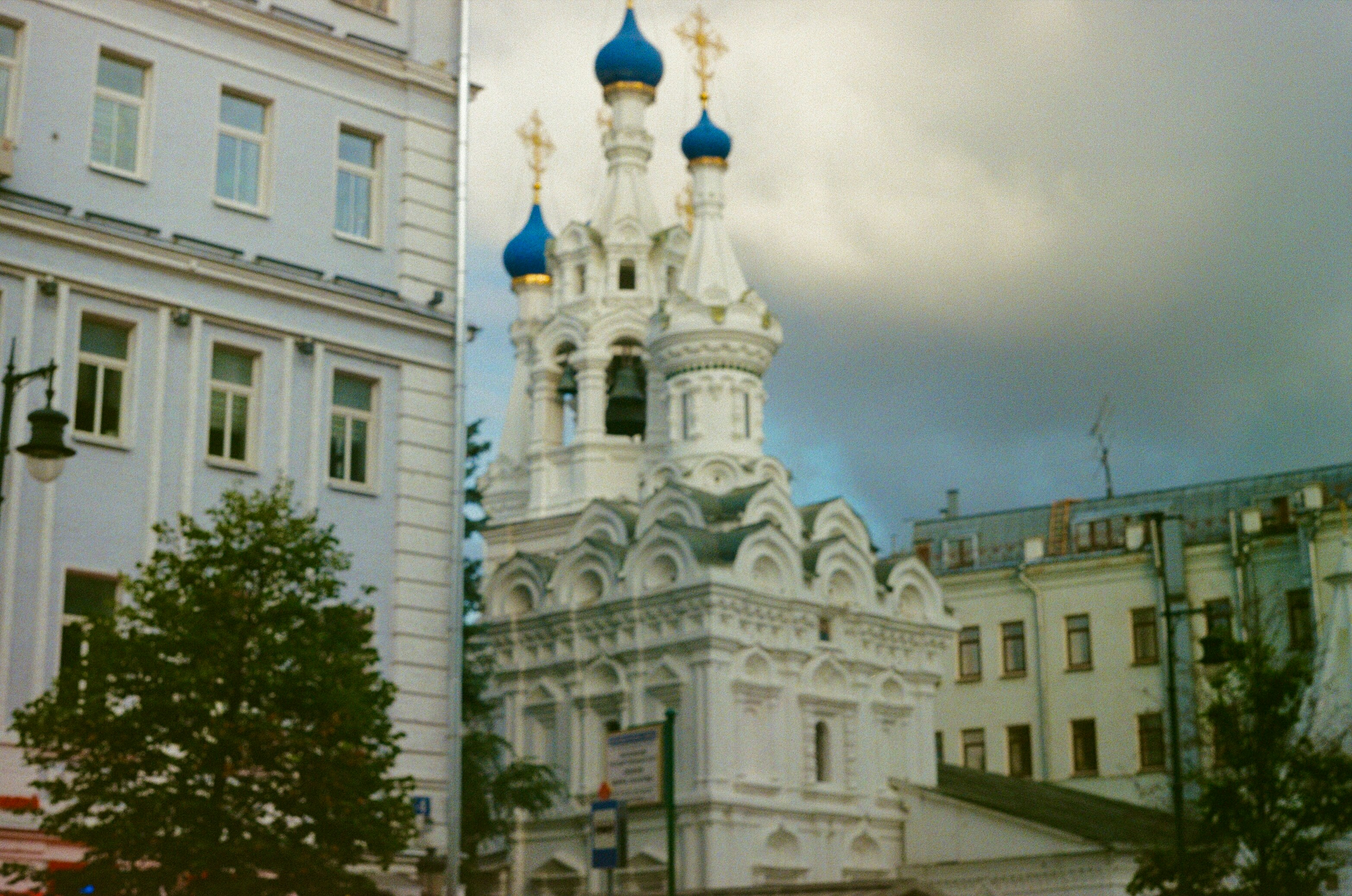 A close‑up, low‑angle film photograph of the Church of the Nativity of the Theotokos in Putinki, one of Moscow's most picturesque 17th‑century churches. The white stone facade and golden domes are captured against a clear evening sky, with part of the building visible from the left. The warm, soft light and analog grain give the scene a nostalgic and majestic atmosphere.