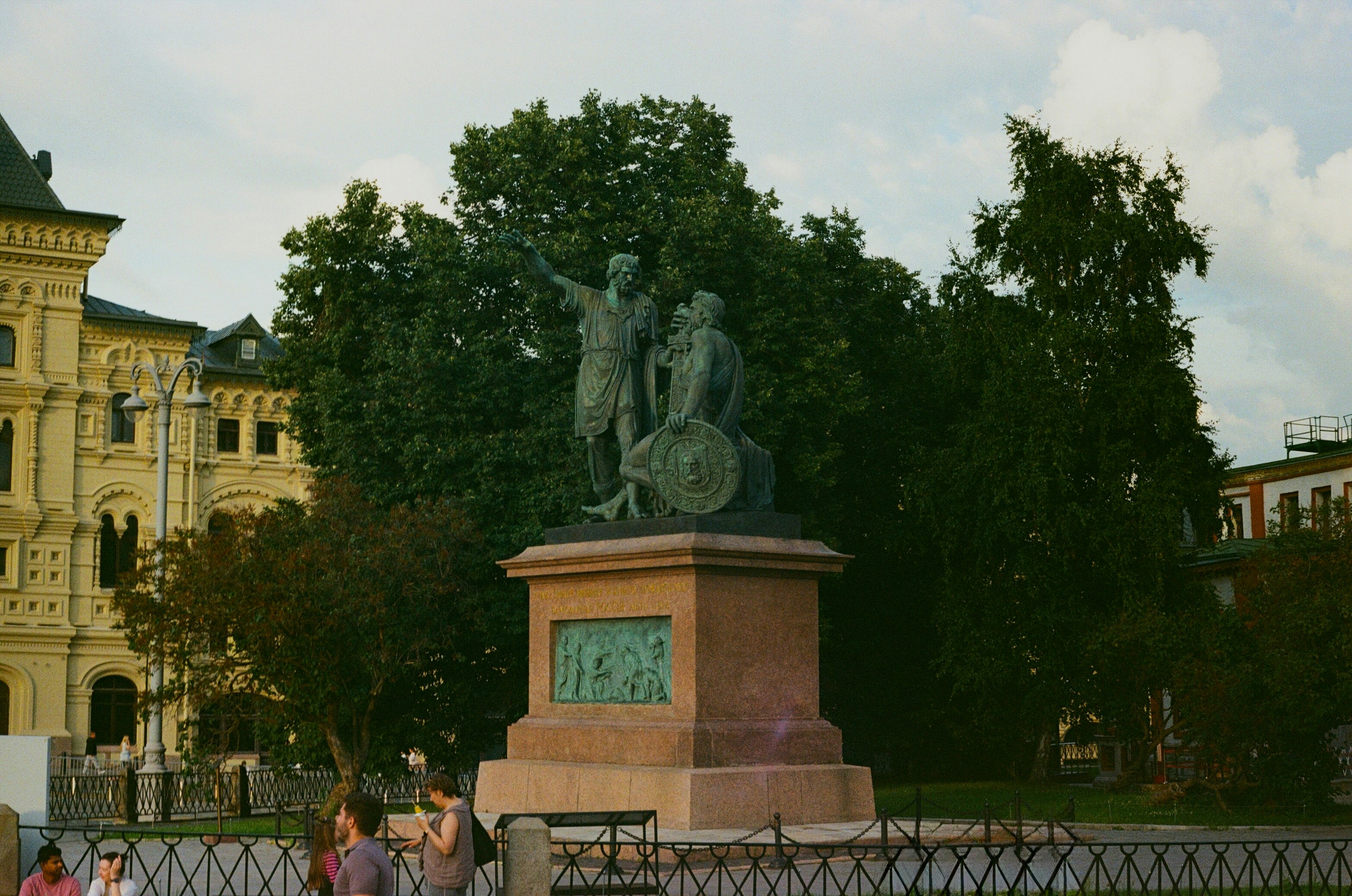 Bronze monument pedestal with two figures in a public square, framed by trees and surrounding architecture.