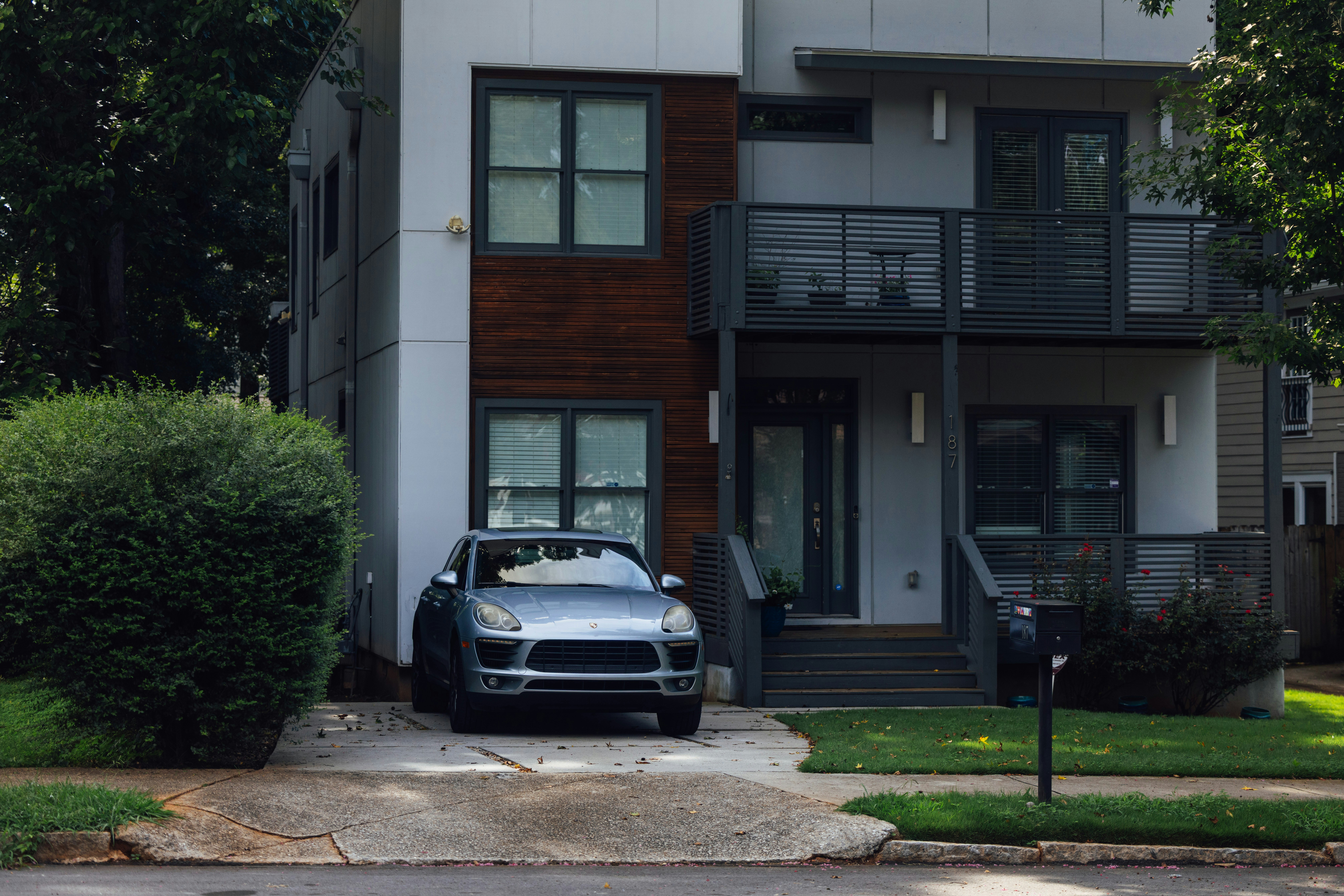 Modern duplex with a silver SUV parked in a concrete driveway, greenery framing a two-level entrance and a small mailbox in the foreground.