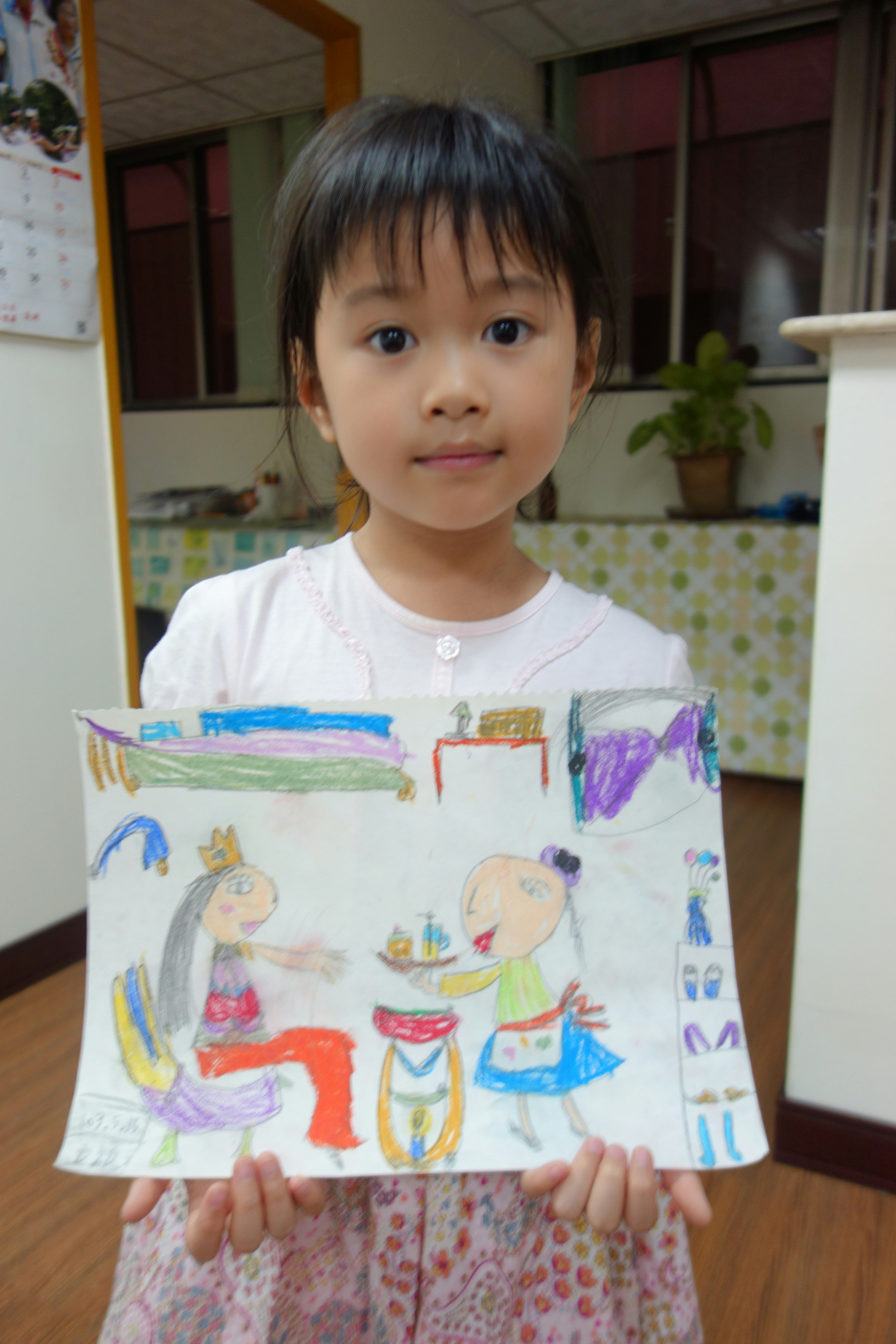 A smiling young girl presents a large drawing, standing indoors with a warm, casual background. This image is a candid portrait featuring a child and her artwork.