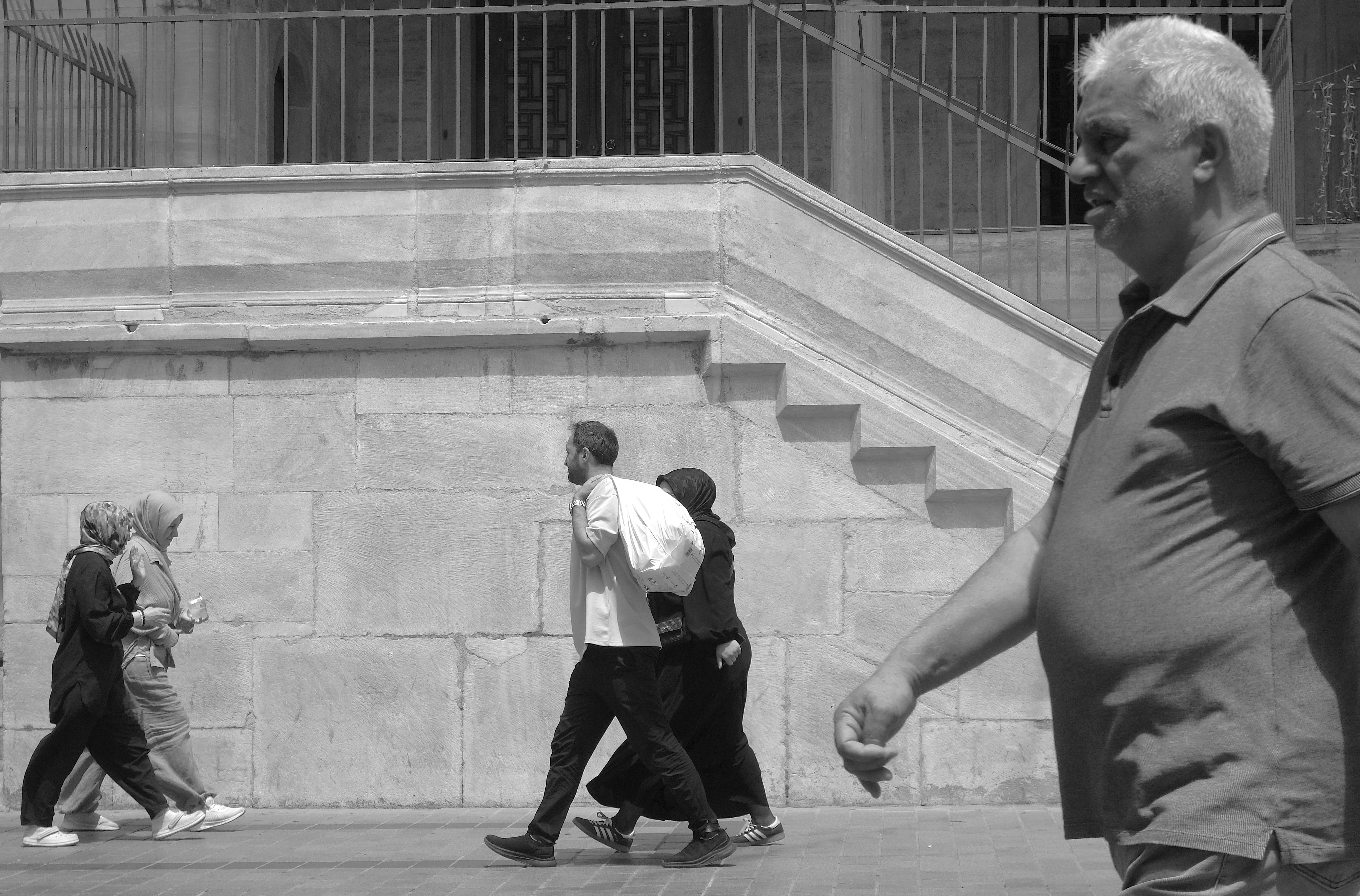 A grayscale street scene with pedestrians walking along a stone wall; a large man in the foreground contrasts with a distant trio, highlighting movement and urban rhythm. This is a photographic capture, not a painting or illustration.